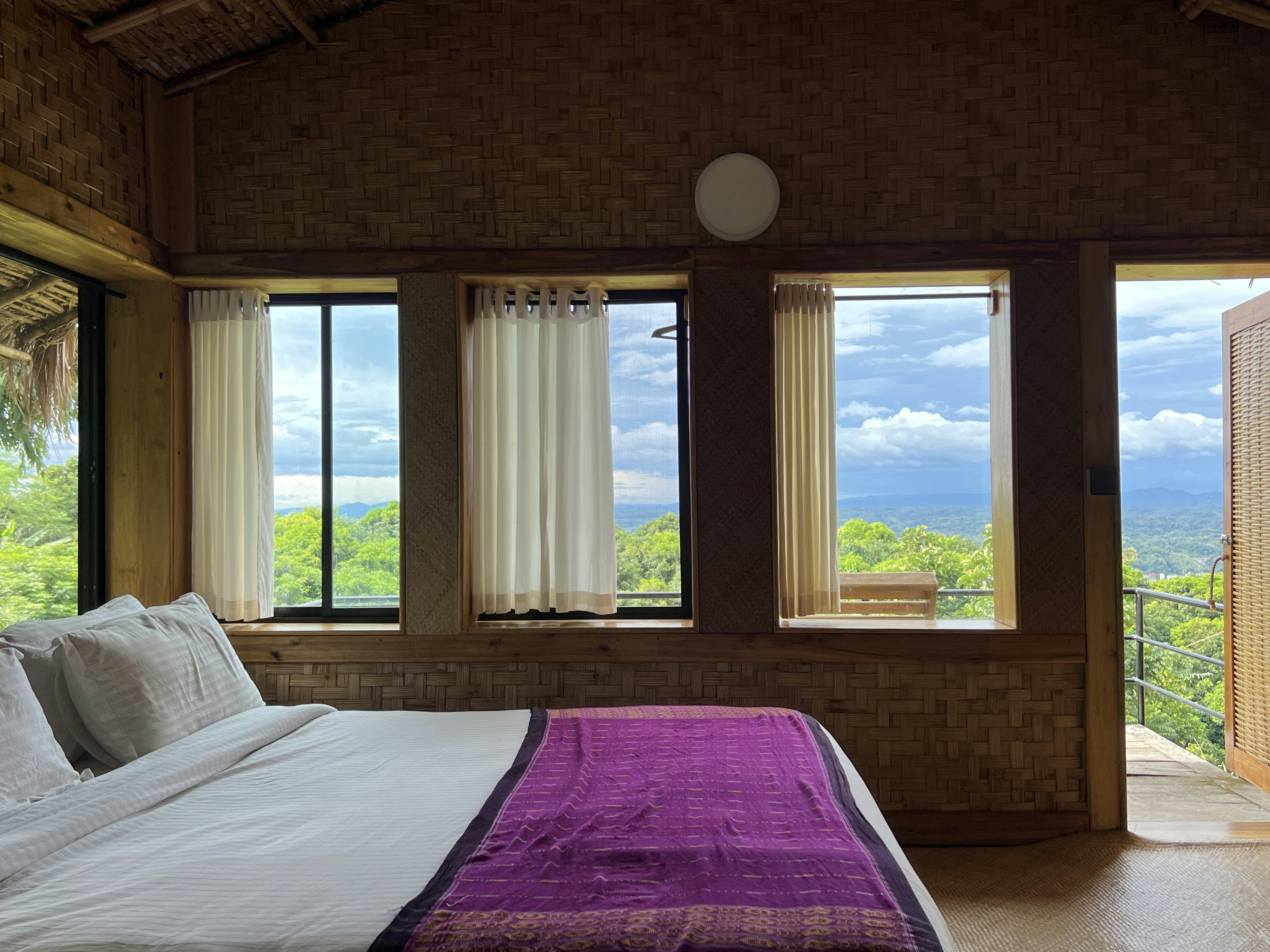 Bright bedroom with large windows revealing a verdant hillside and distant sea. A white bed dominates the foreground with a bold purple runner against warm wood walls.