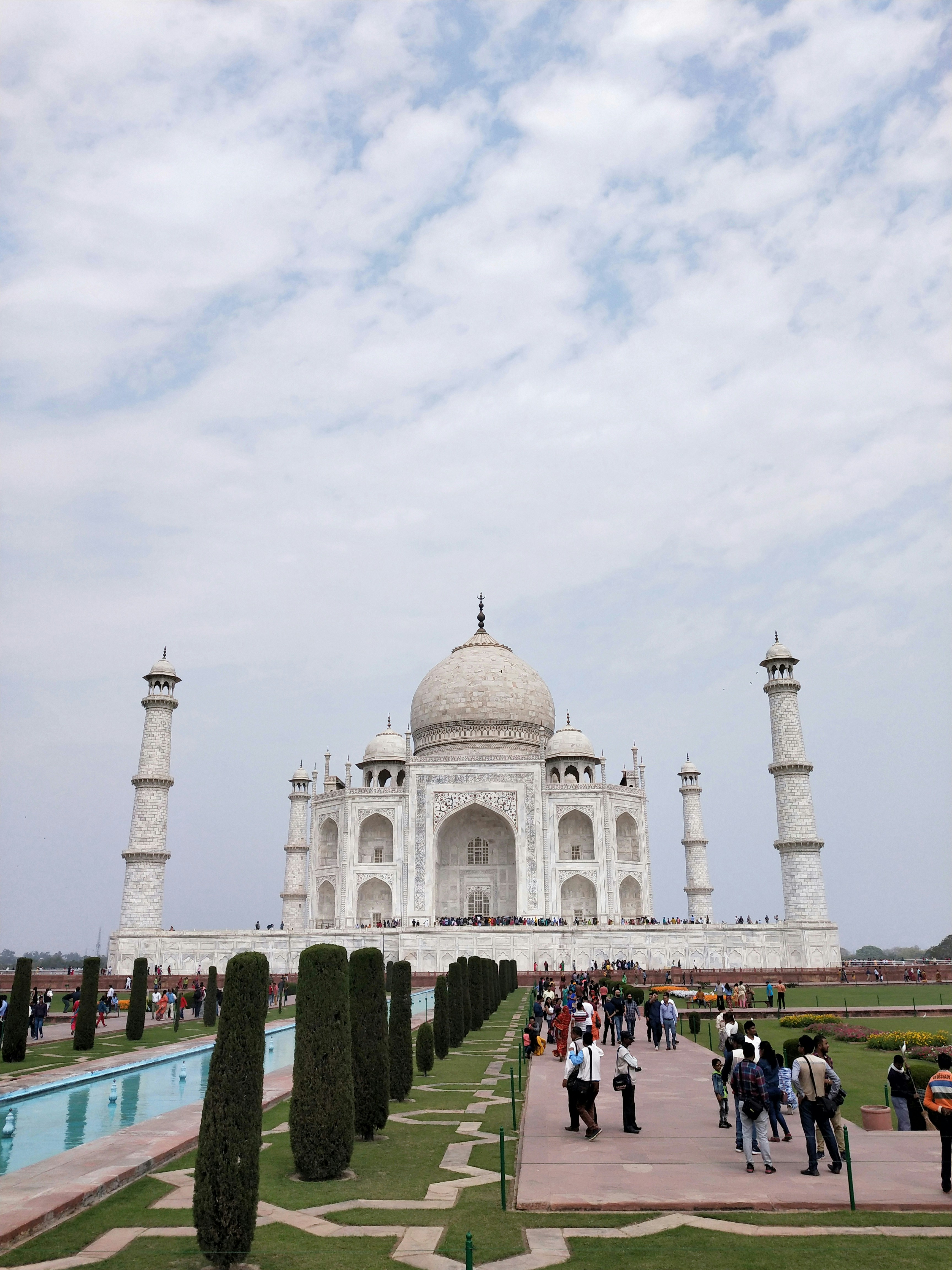 The Taj Mahal stands grandly against a cloudy sky, surrounded by lush gardens and visitors exploring its beauty.