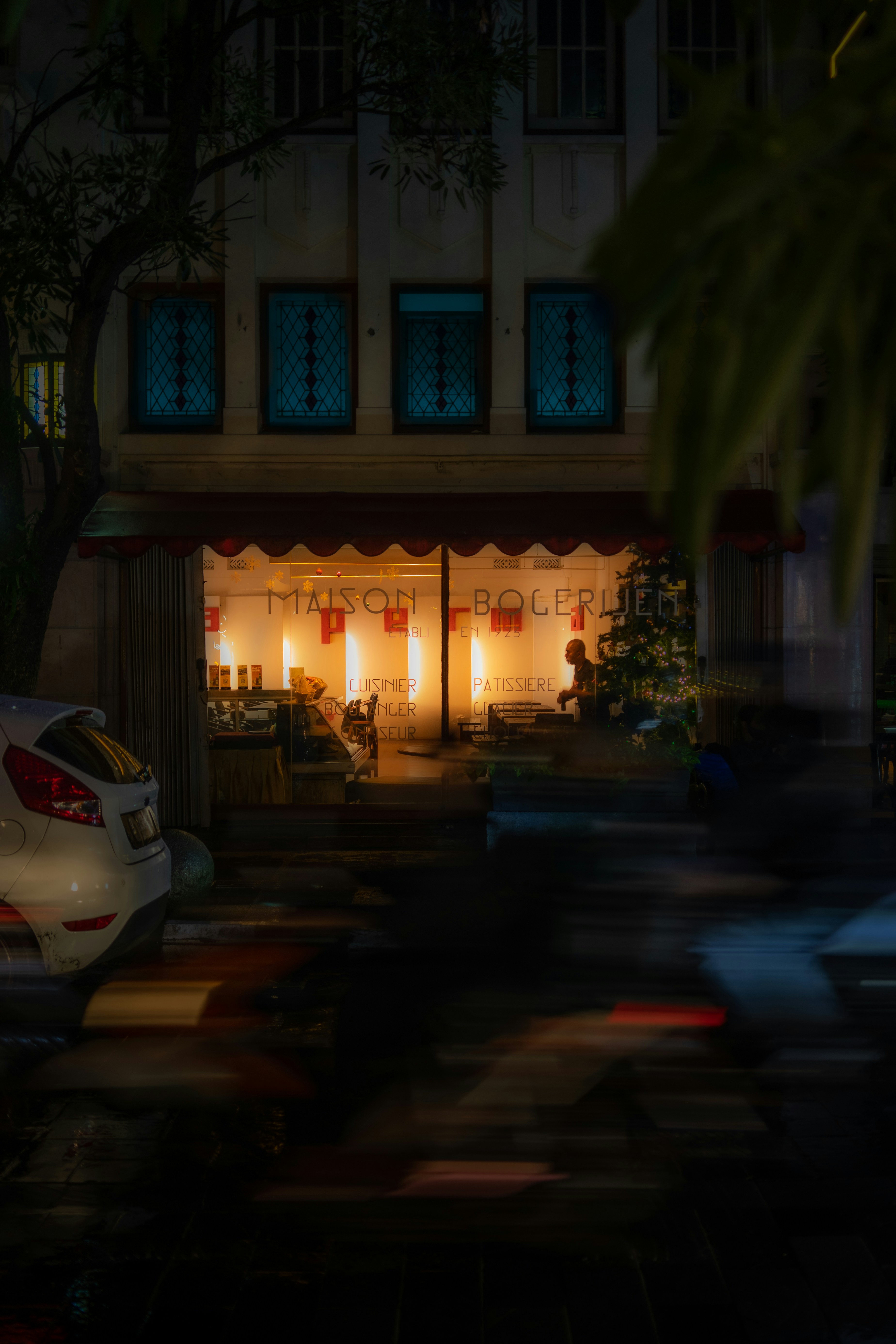Warmly lit bakery storefront inviting passersby with its cozy atmosphere and tantalizing treats. The blurred motion of vehicles adds a dynamic urban feel.