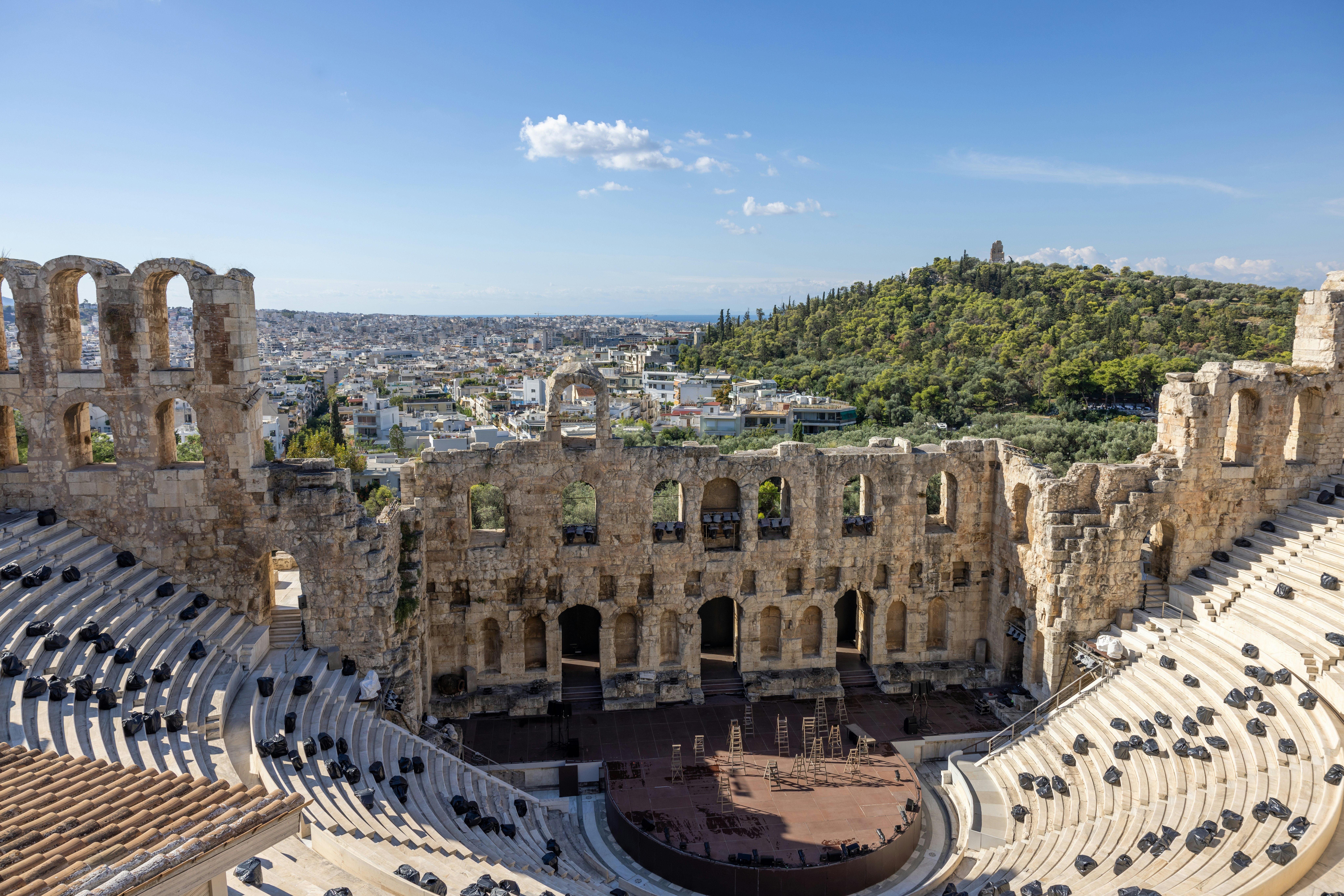 Ancient amphitheater overlooking a sprawling cityscape with a lush green hill in the background under a clear blue sky.