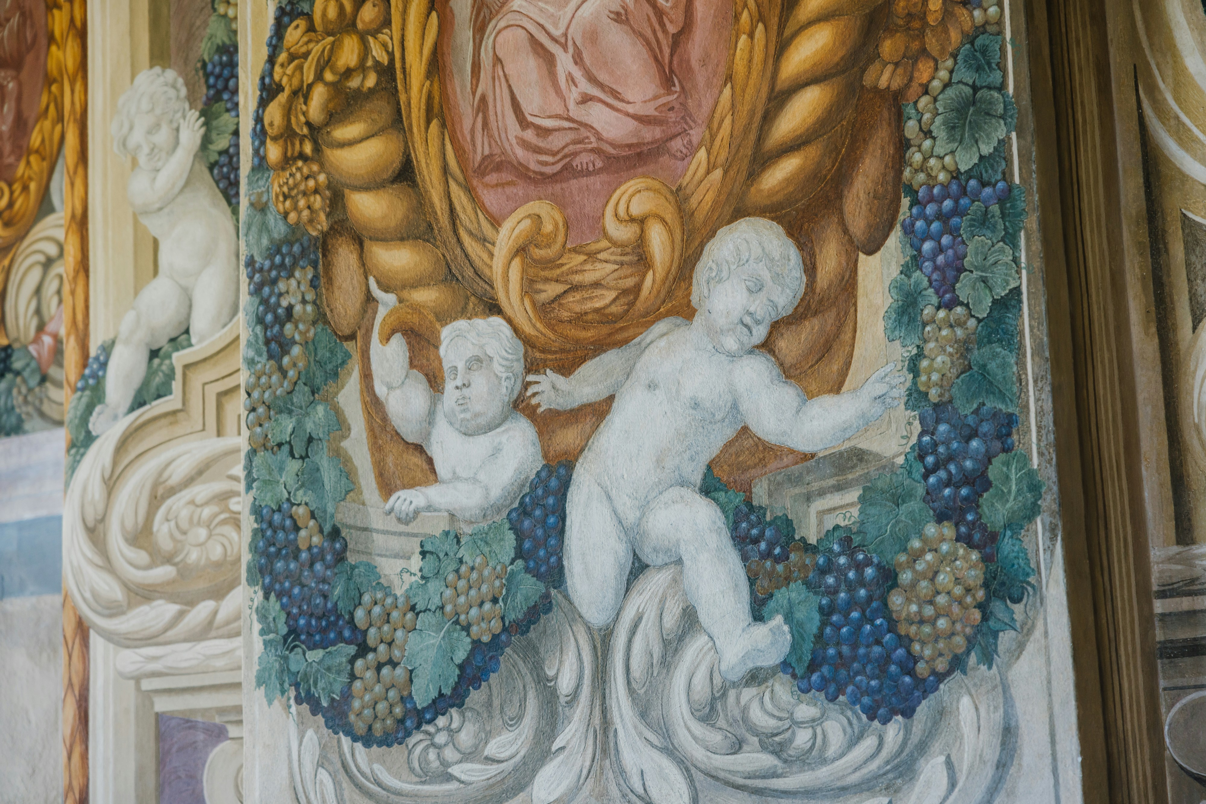 Two cherubic figures playfully interact among intricate grapevine motifs in a classic fresco. The artwork showcases the harmony of nature and artistry.