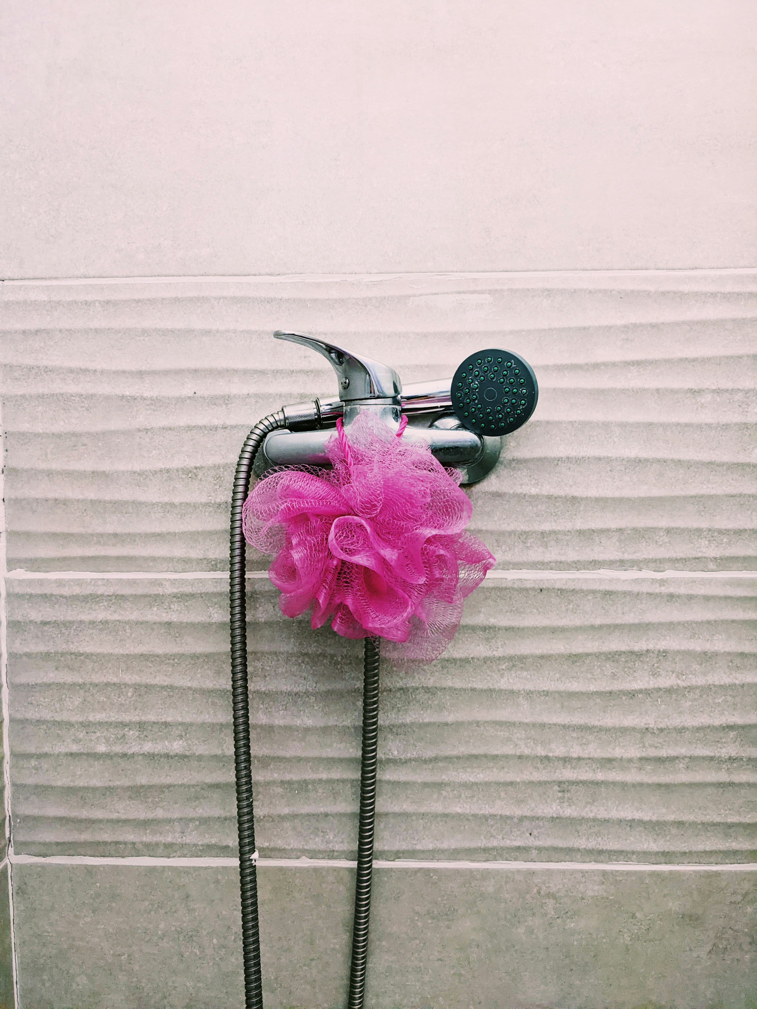 Shower faucet with a pink loofah hanging from the spout against textured, light-colored tiles.