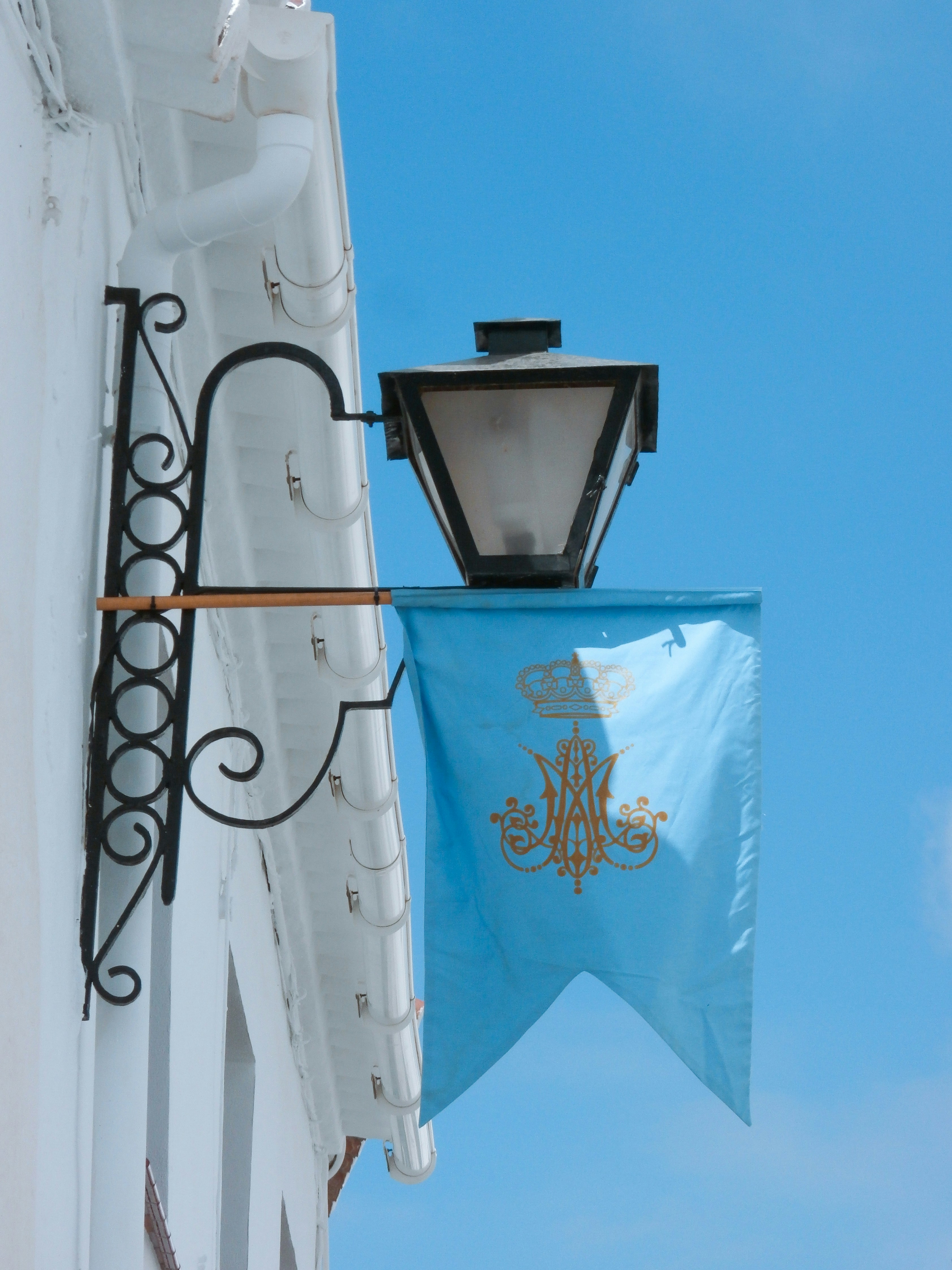 A black wrought iron wall-mounted street lamp is attached to a white building with visible roof gutters. Below the lamp, a light blue banner with a gold monogram and crown emblem hangs vertically. The background shows a clear blue sky. The photo was taken in Mijas, Spain.