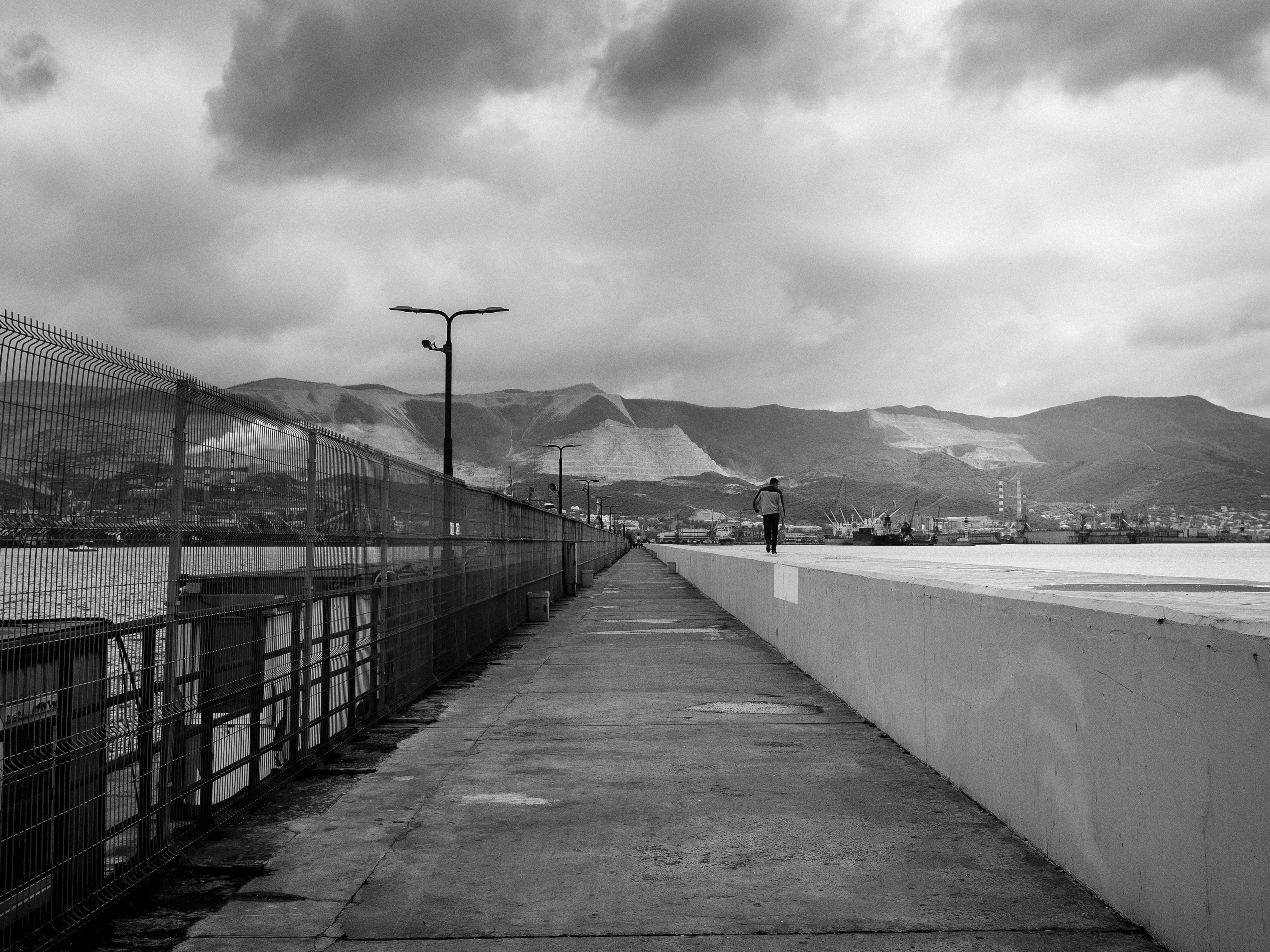 Black and white view of a concrete pier leading to distant mountains under dramatic clouds.