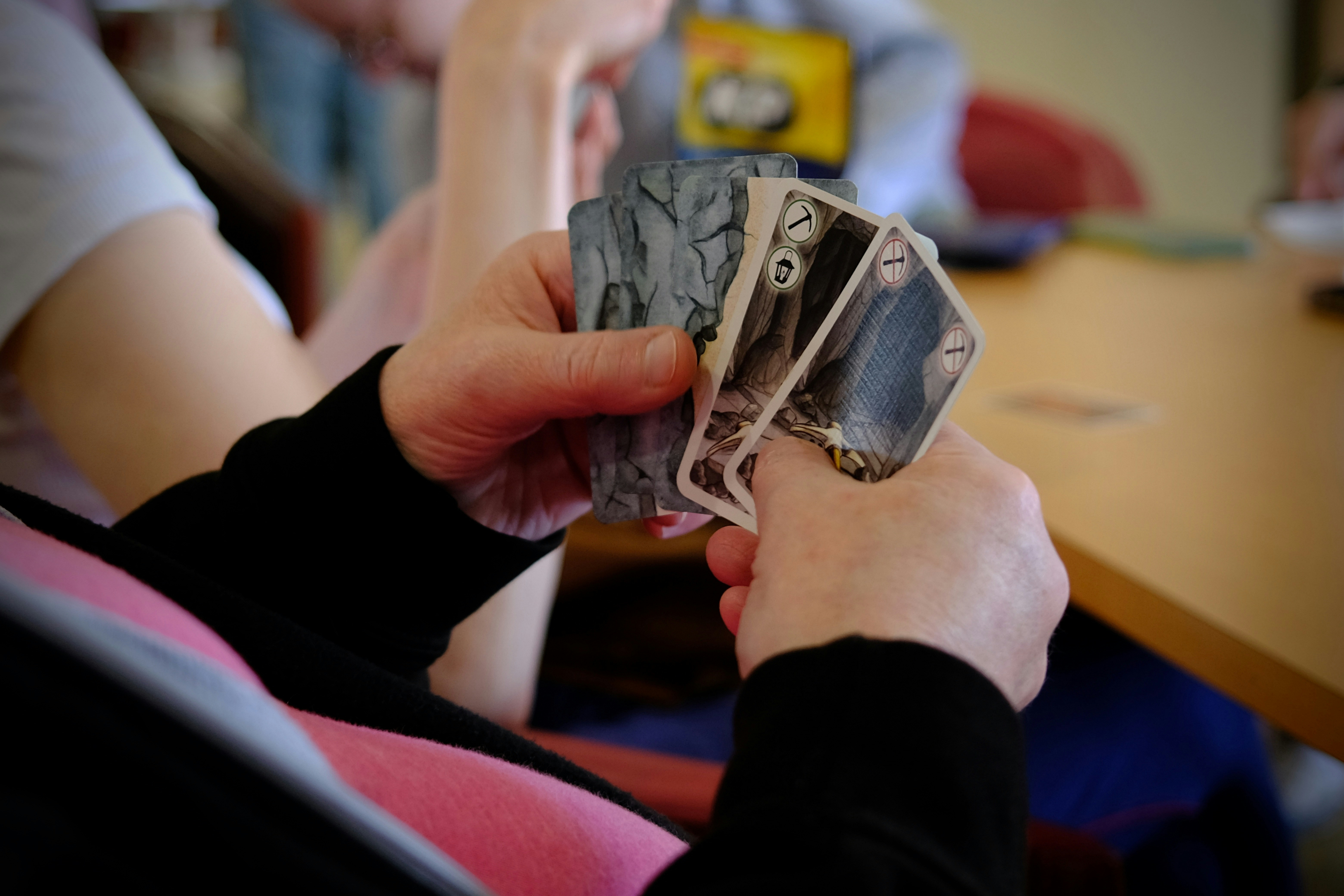 Hands holding a set of unique playing cards during a game, with blurred figures in the background. The focus is on the intricate designs of the cards.