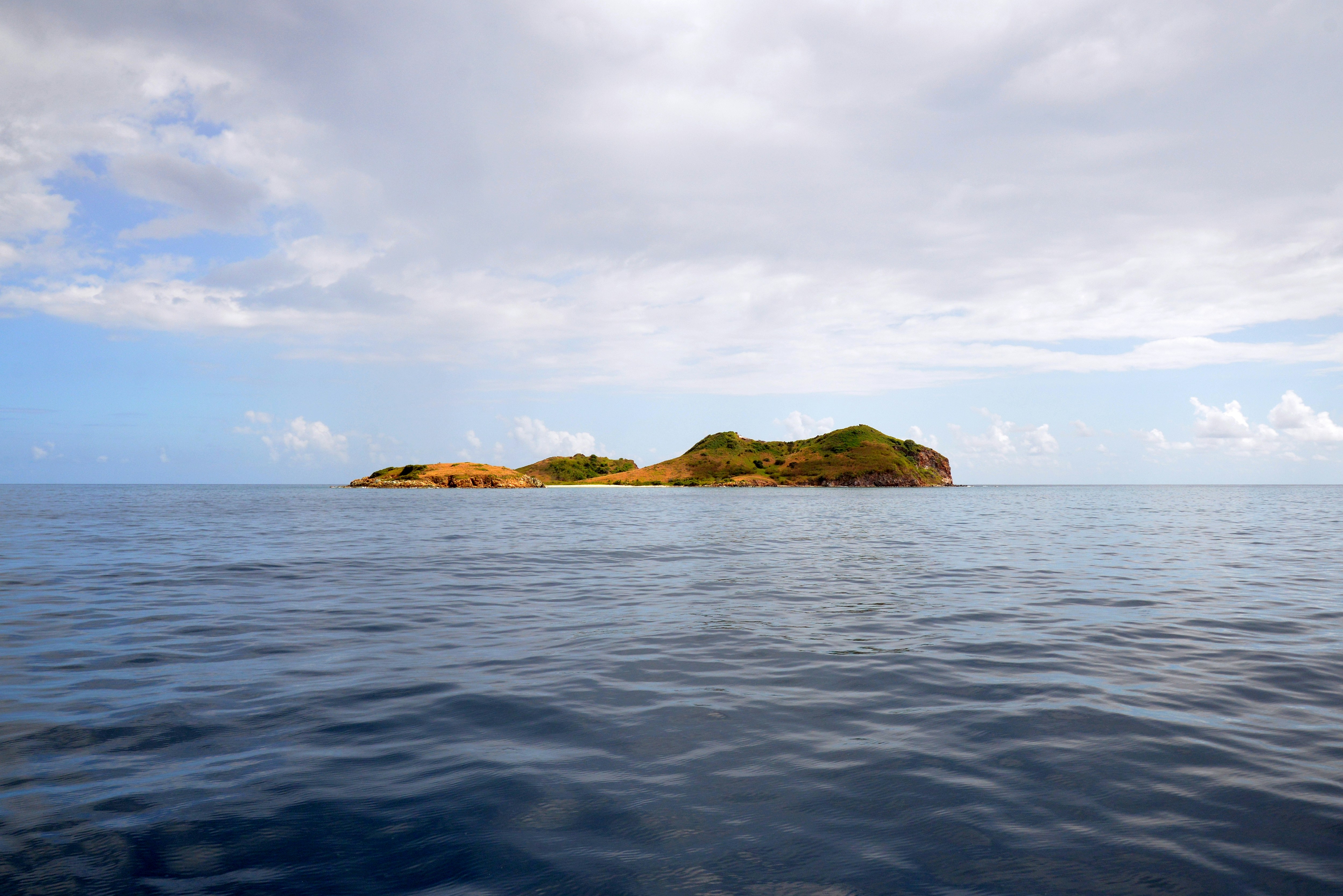 A tranquil island rises from the calm sea under a partly cloudy sky, showcasing lush greenery and rocky shores.