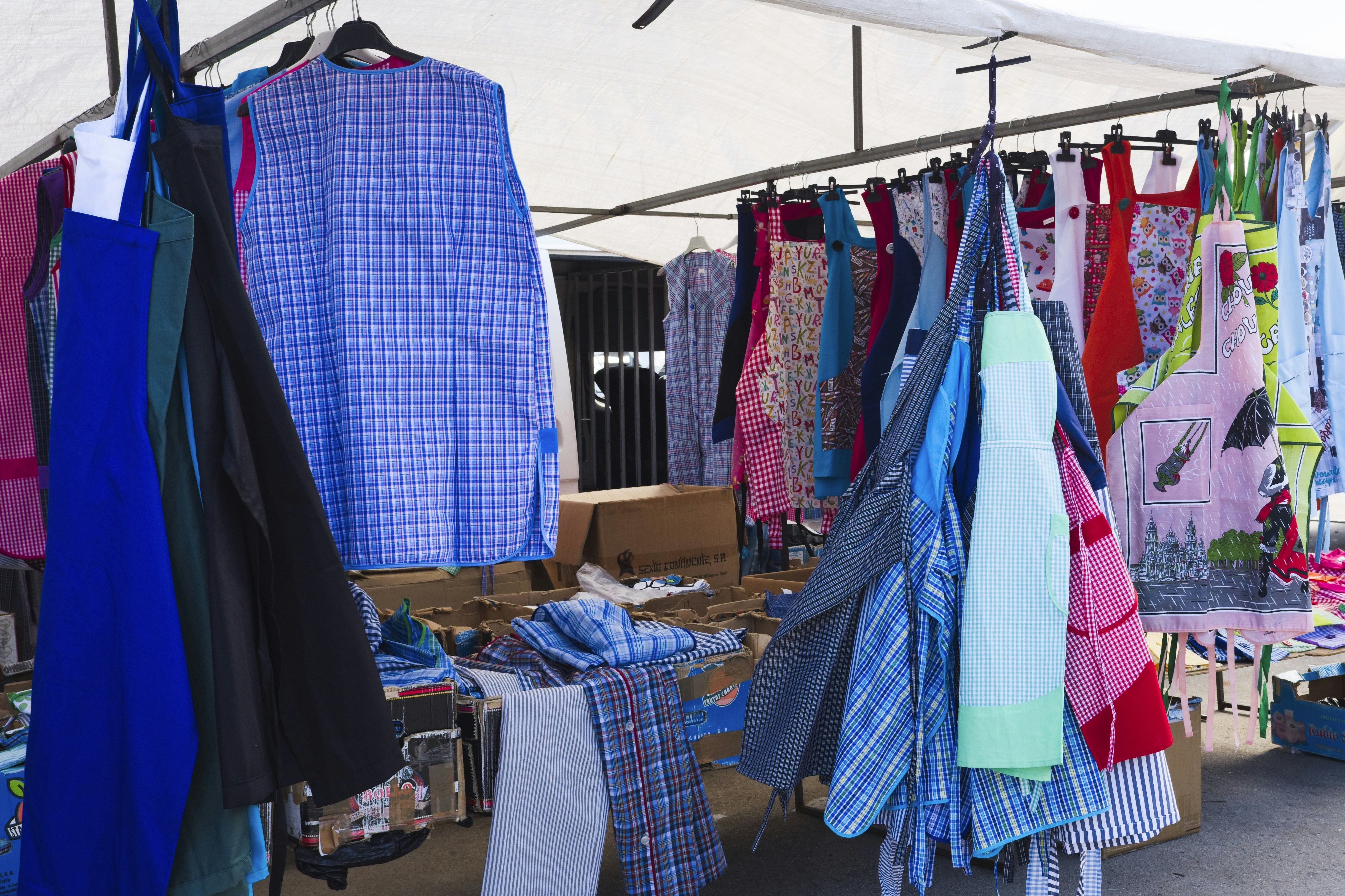 Colorful Aprons Hanging For Sale At Outdoor Market - A variety of colorful aprons are hanging for sale on a rack under a white canopy at an outdoor market. The aprons are made of different fabrics and patterns, including checkered, floral, and solid colors. Some of the aprons have pockets, while others have ties.