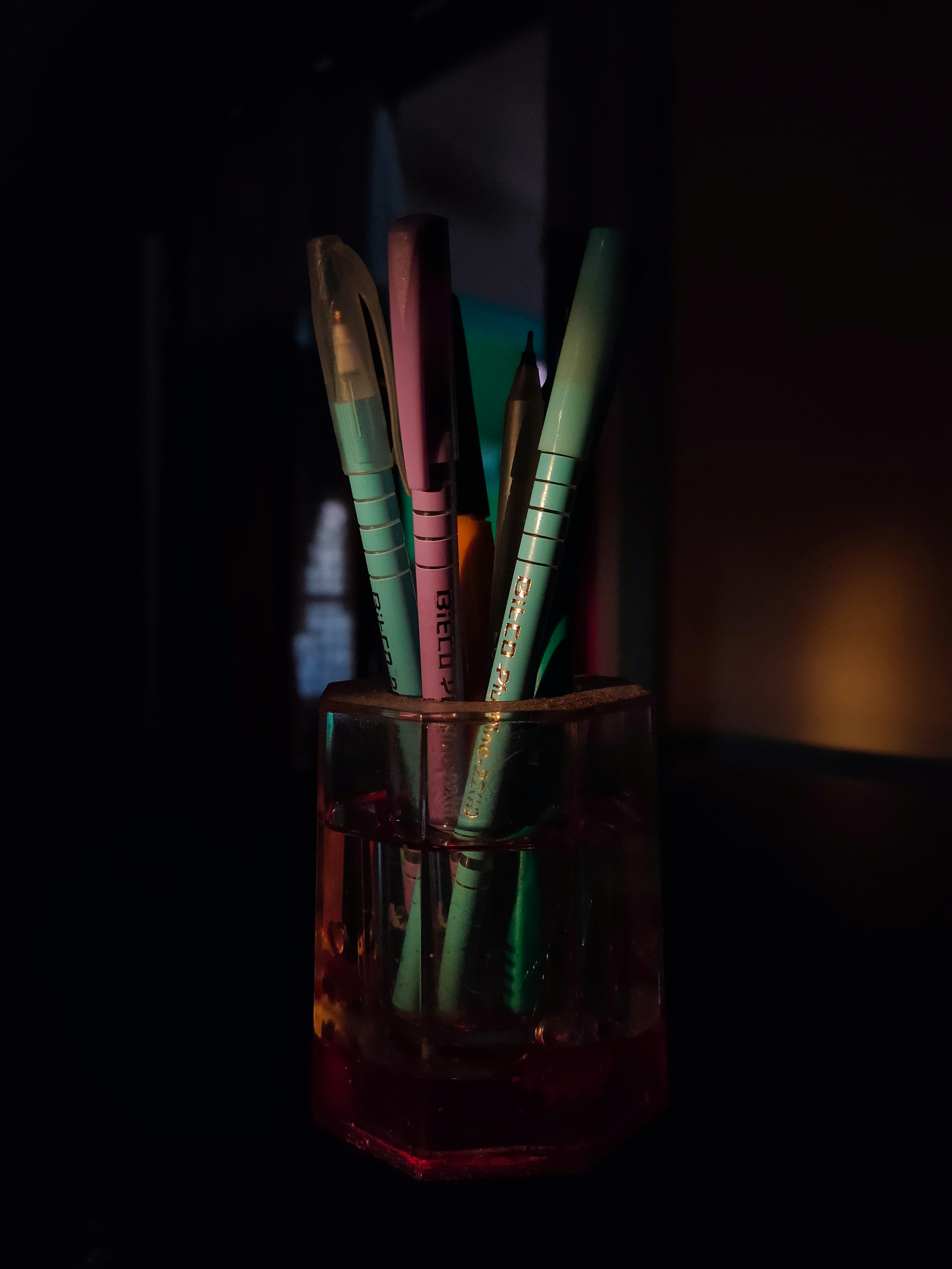 A collection of colorful pens arranged in a glass container, illuminated by soft light, creating a contrast between vibrant colors and dark surroundings.