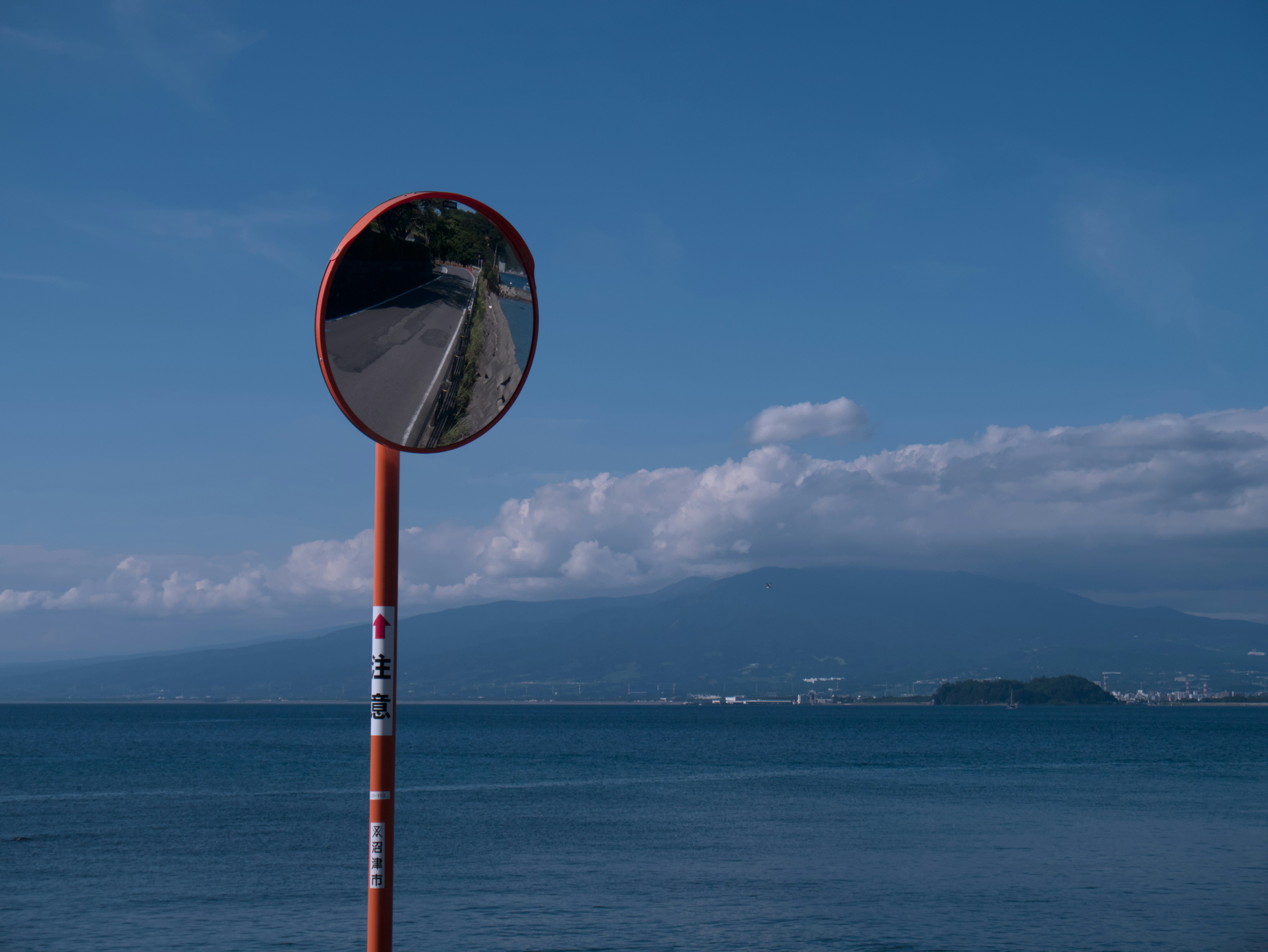 Round convex safety mirror mounted on a red-and-white striped pole reflects a glimpse of the road. The scene sits by a calm sea beneath a clear blue sky.