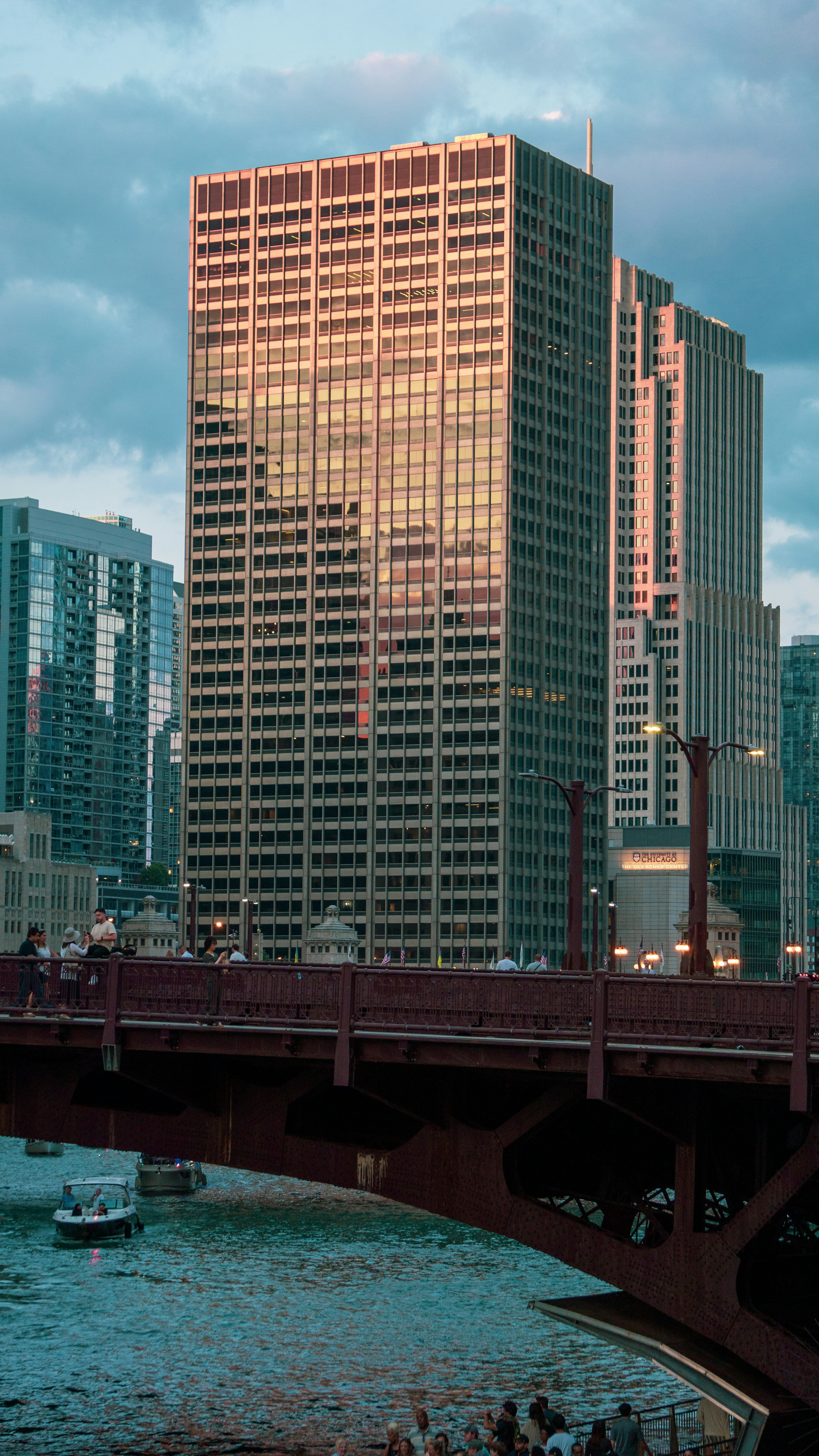 Modern skyscrapers reflecting the evening light over a bustling river, with a bridge and pedestrians below.
