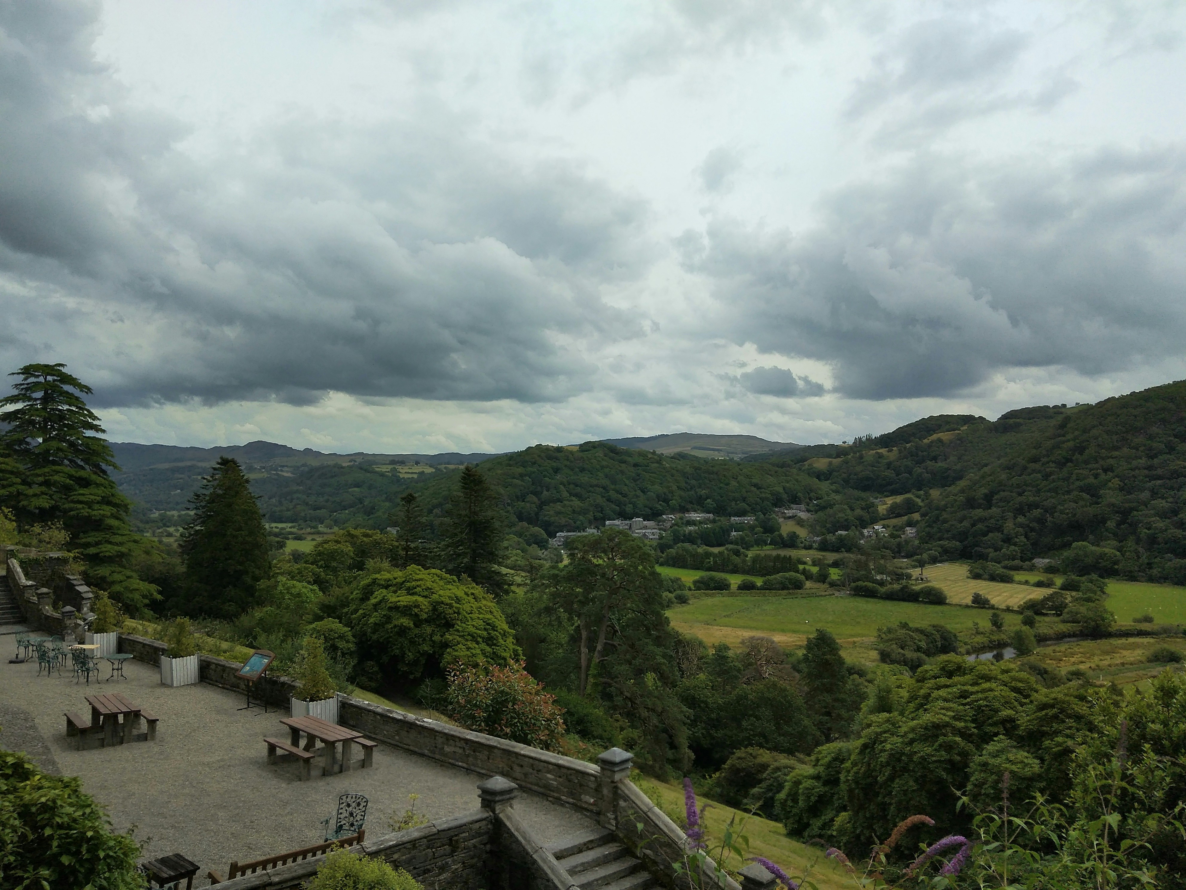Lush green hills and valleys stretch beneath a dramatic sky filled with clouds, showcasing the serene beauty of the landscape. The scene captures a tranquil moment in nature.