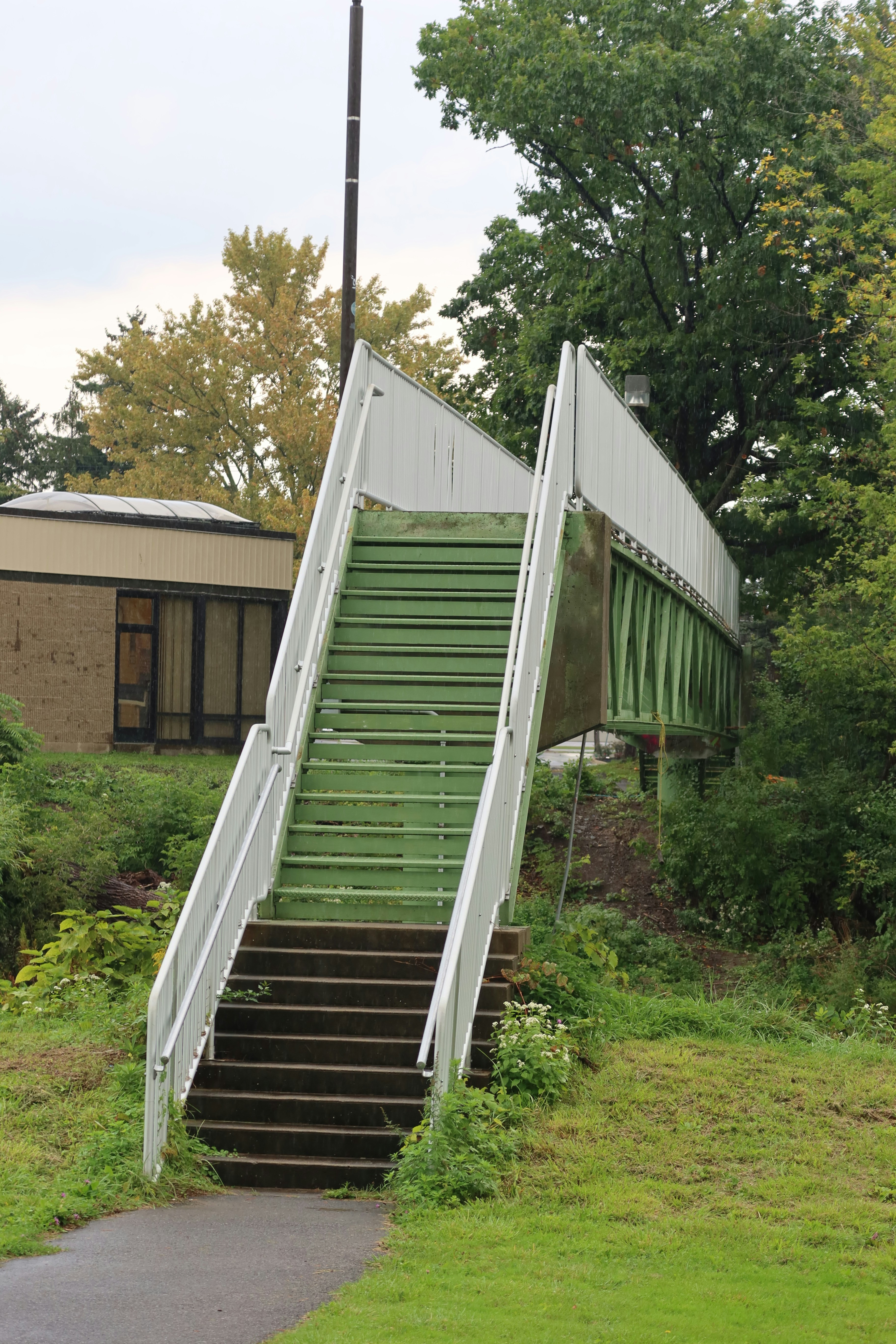 Photograph of a metal-framed footbridge staircase with green steps spanning a small park, backed by a brick building and lush trees.