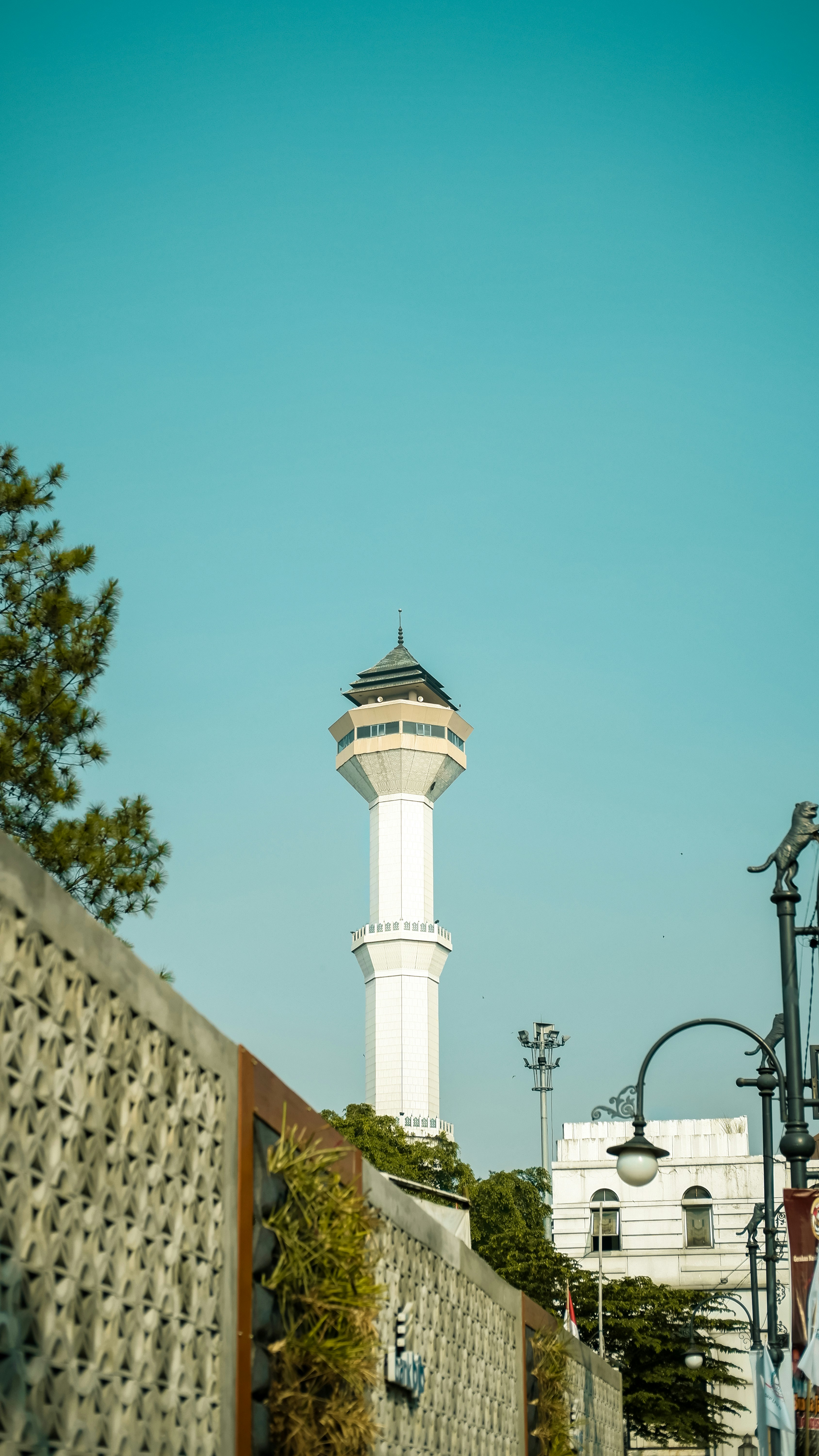 A modern observation tower stands tall against a clear blue sky, framed by greenery and urban architecture. The scene captures the blend of nature and city life.