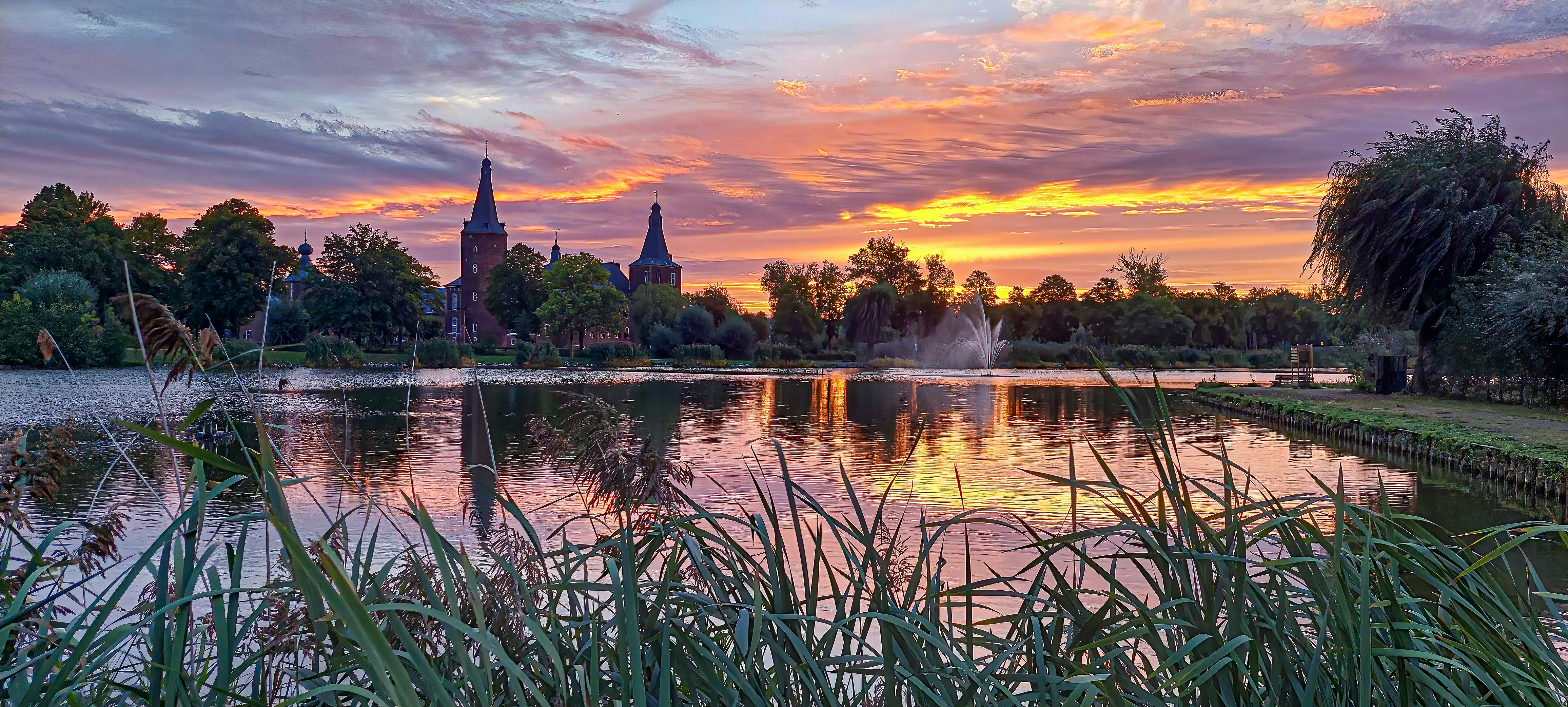 Sunset over a calm lake with a central fountain, reflected light shimmering on the water, and tall foreground grasses. Distant trees and spire silhouettes frame the scene.