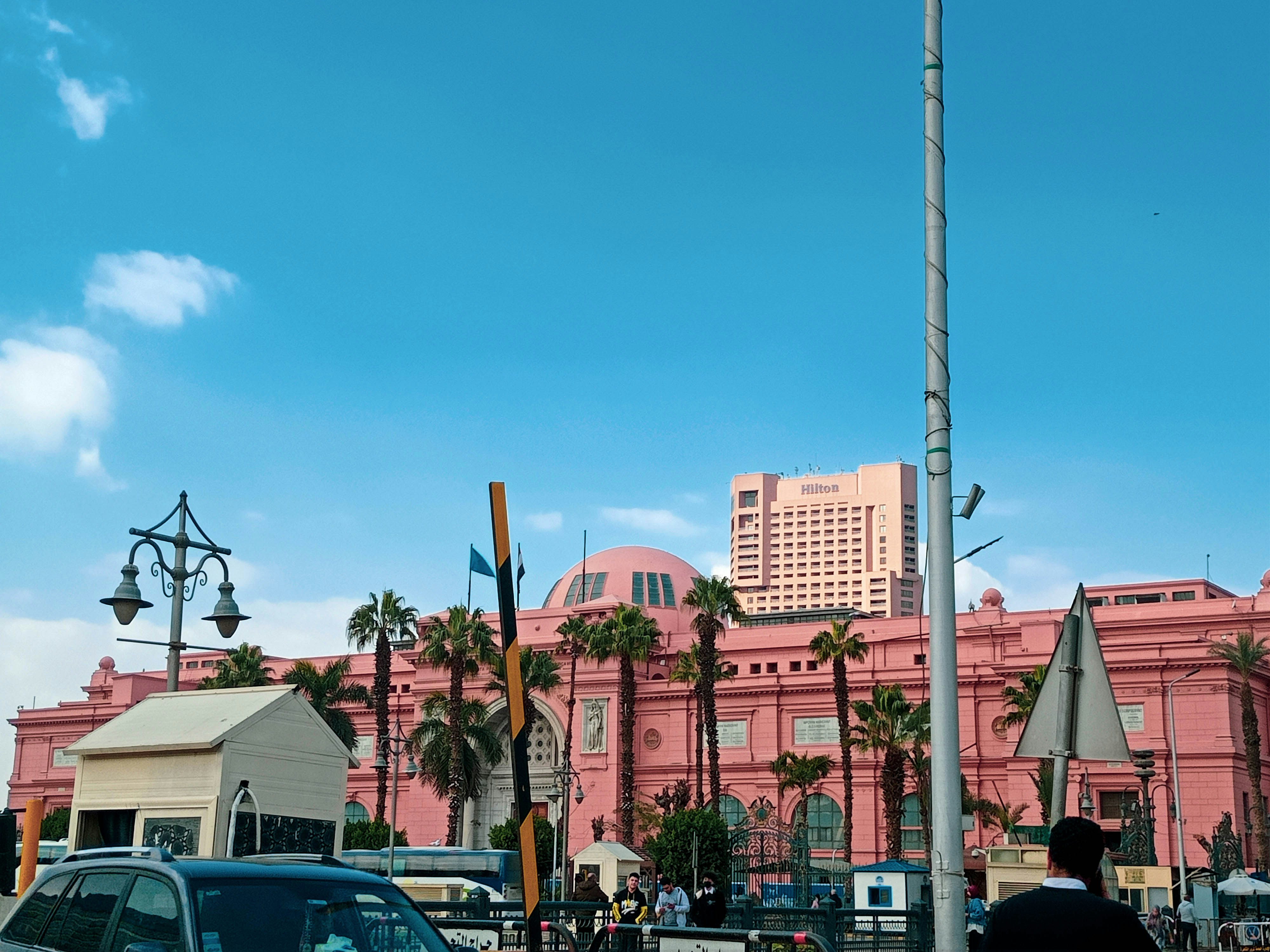 Pink building with palm trees and a flagpole under a bright blue sky.
