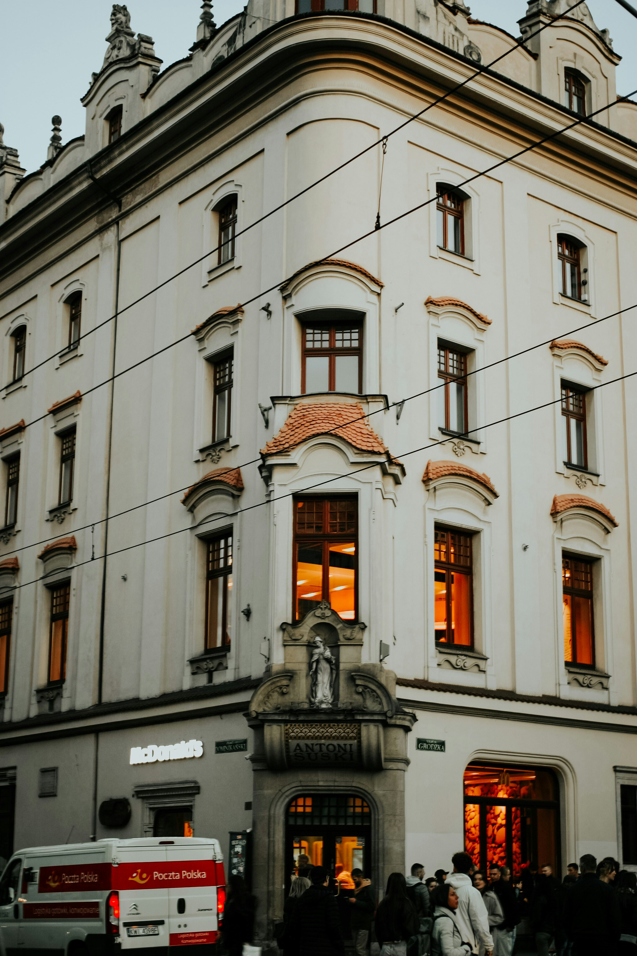 Ornate white building facade with arched windows glowing orange from interior lights, while pedestrians crowd the entrance beneath a carved arch.