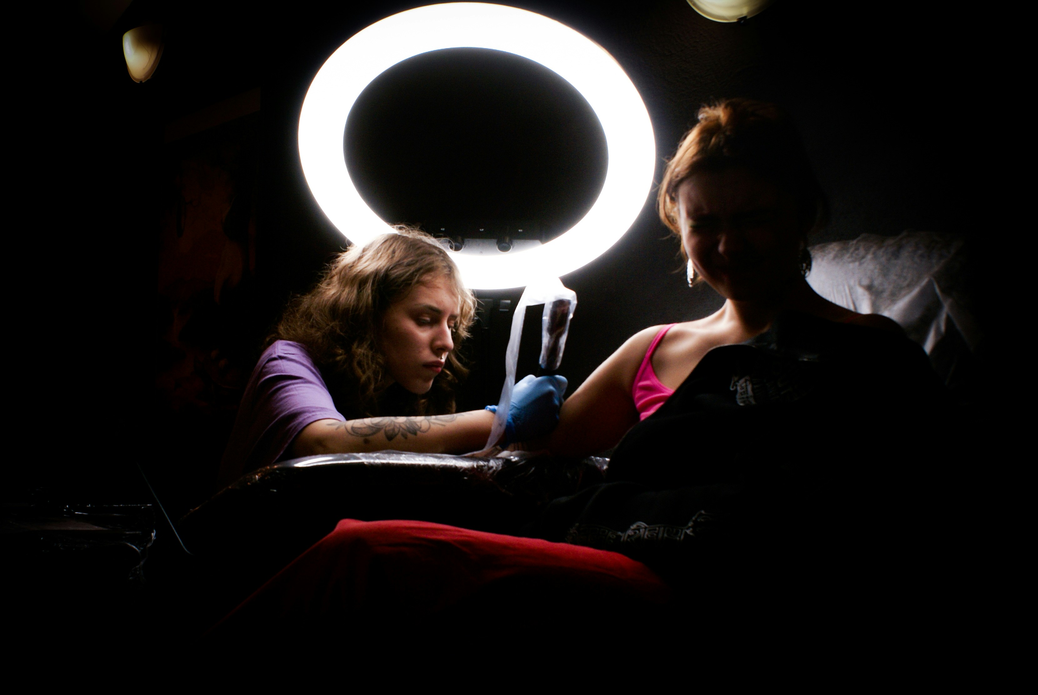 Two people work on a tattoo session under a circular ring light in a dark studio. The tattoo artist focuses on the task as a client sits nearby in shadow.
