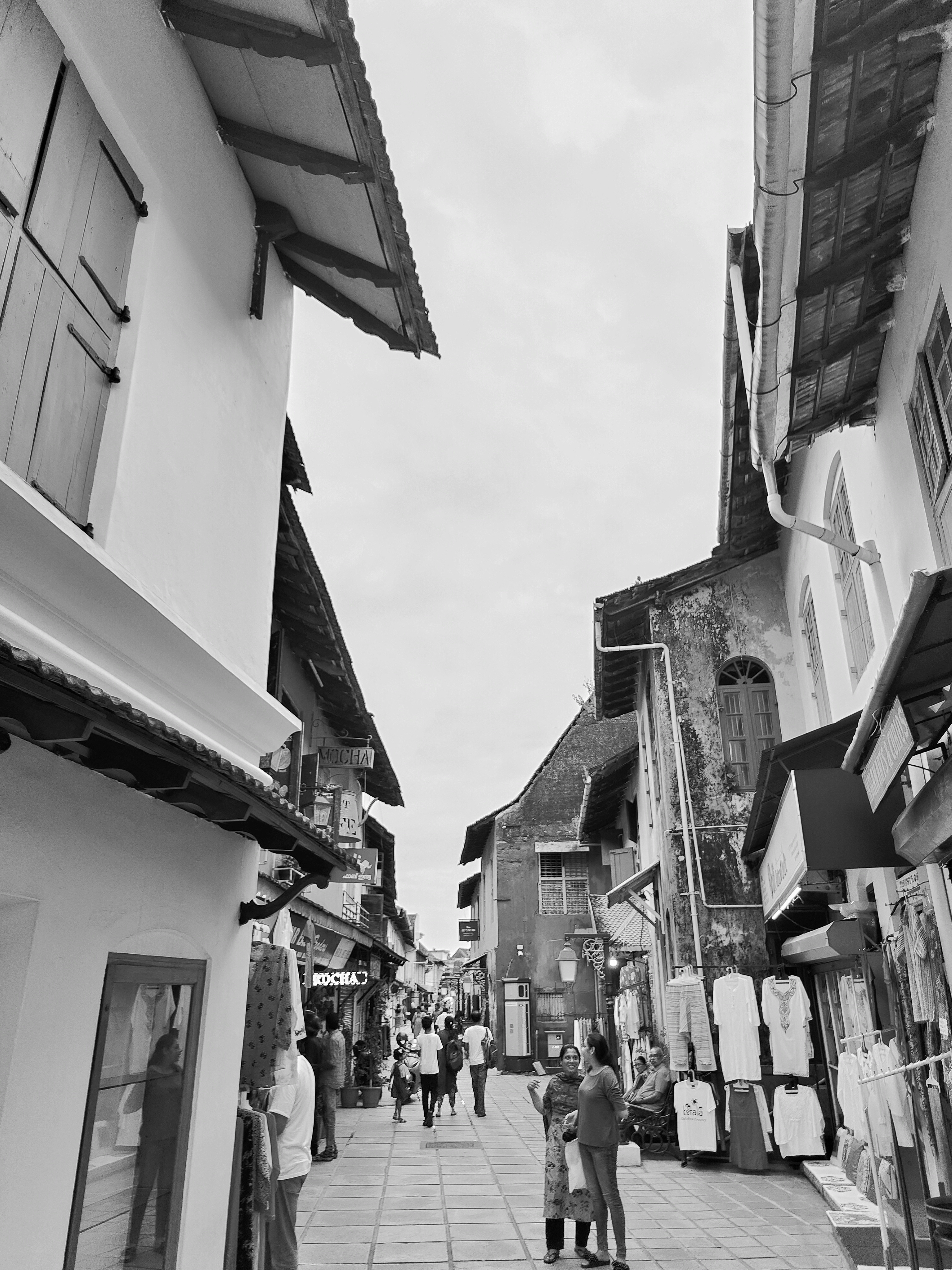 Photograph of a narrow pedestrian alley lined with shopfronts and people walking, rendered in black and white.