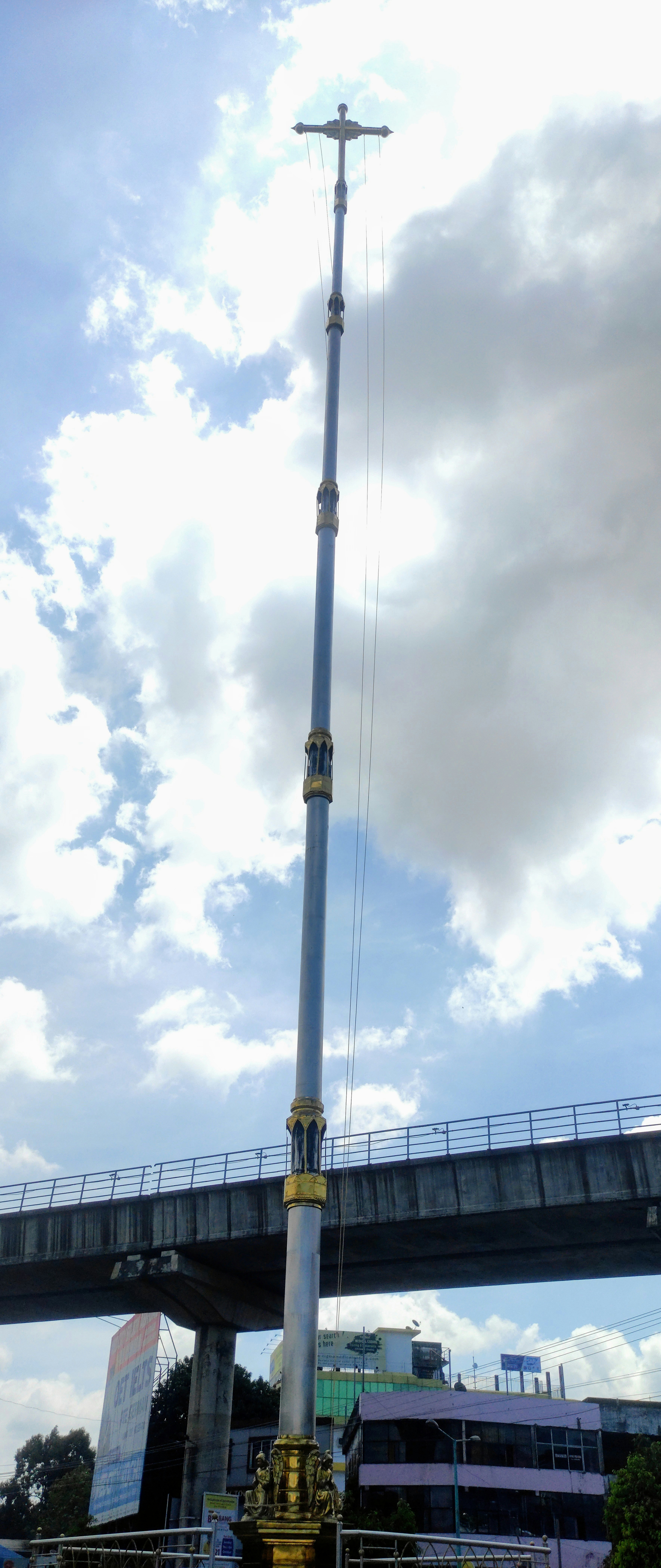 A tall decorative pole stands against a backdrop of dynamic clouds, showcasing intricate details at its base and a cross atop. The scene captures an architectural landmark amidst urban elements.