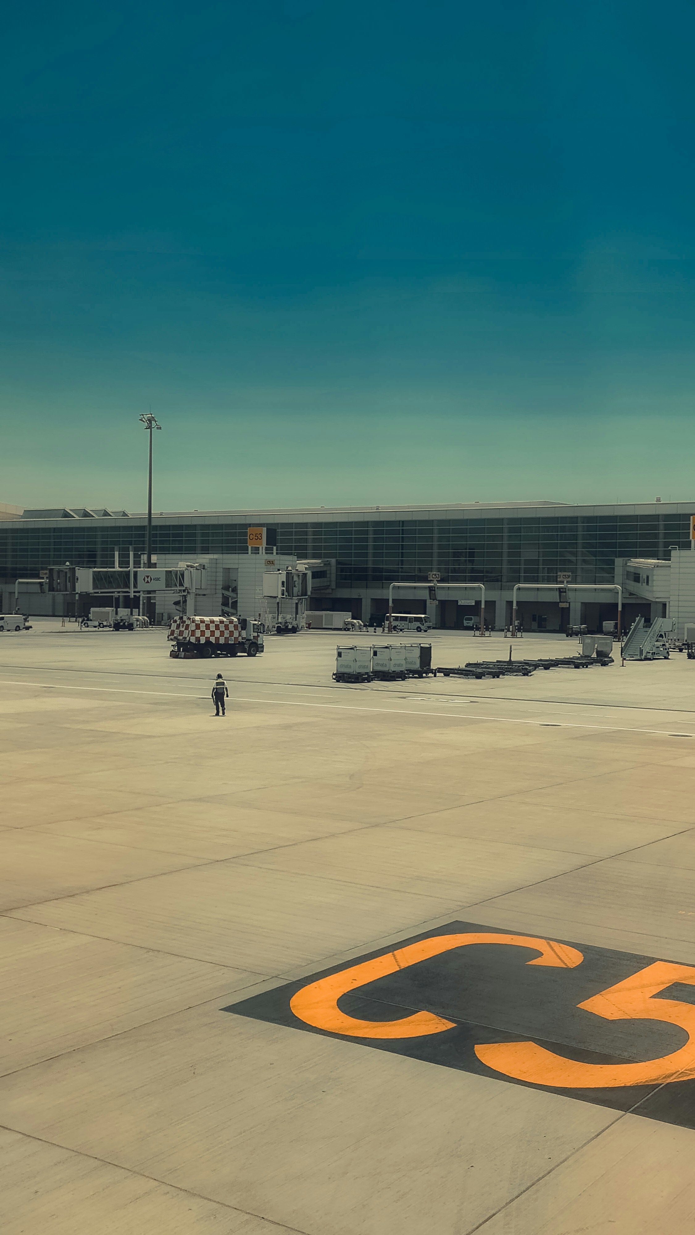 Ground crew member walking across the airport tarmac near terminal gates, with cargo and service vehicles in the background.