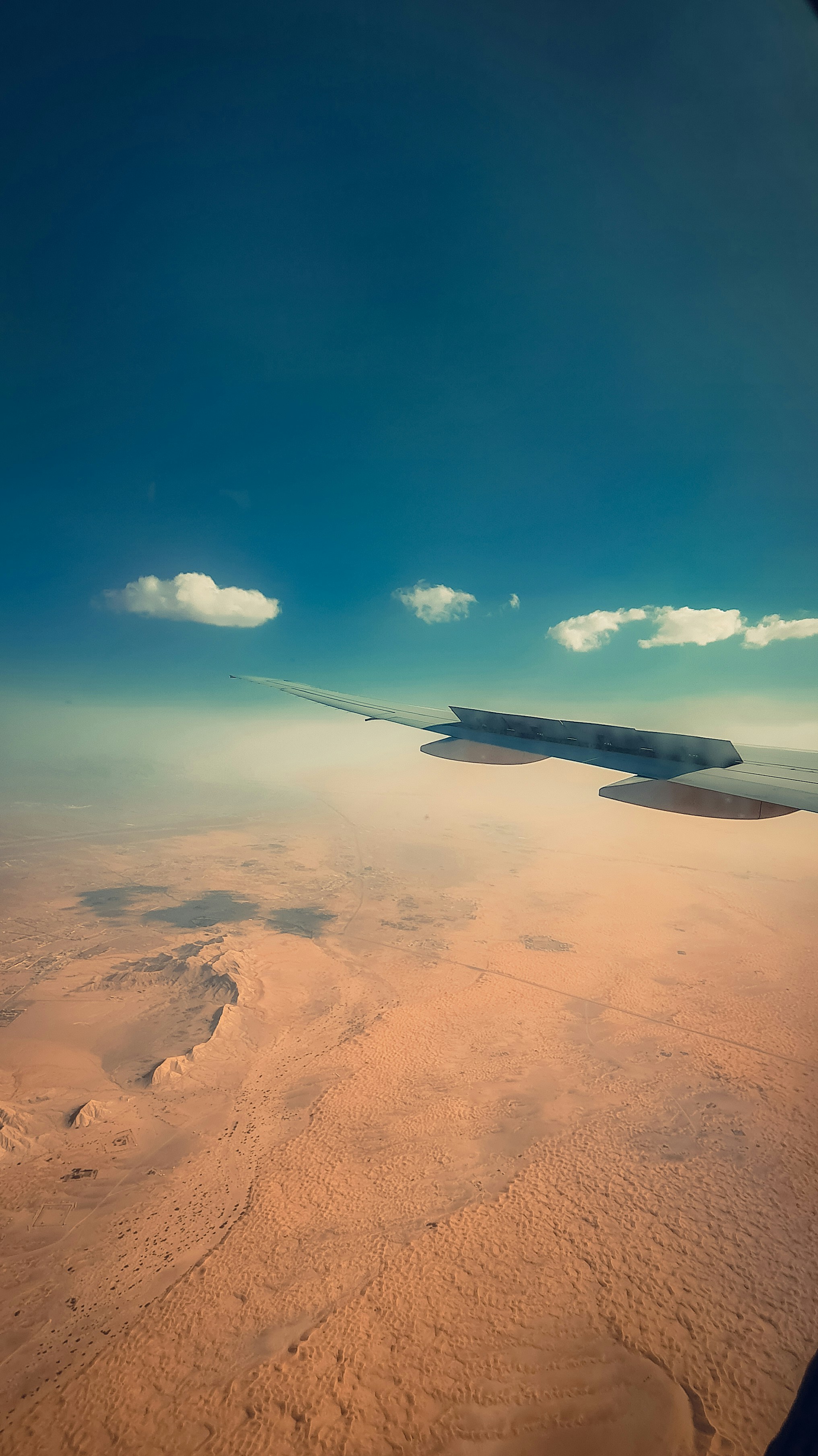 Aerial photograph of a plane wing extending into a gradient blue sky, with vast desert terrain and a few clouds below.