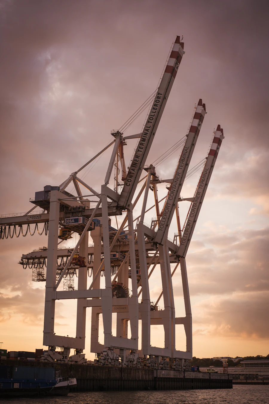 Port cranes rise over the quay at sunset, their forms bathed in warm light against a cloud-streaked sky. The composition highlights towering steel against the harbor's quiet horizon.