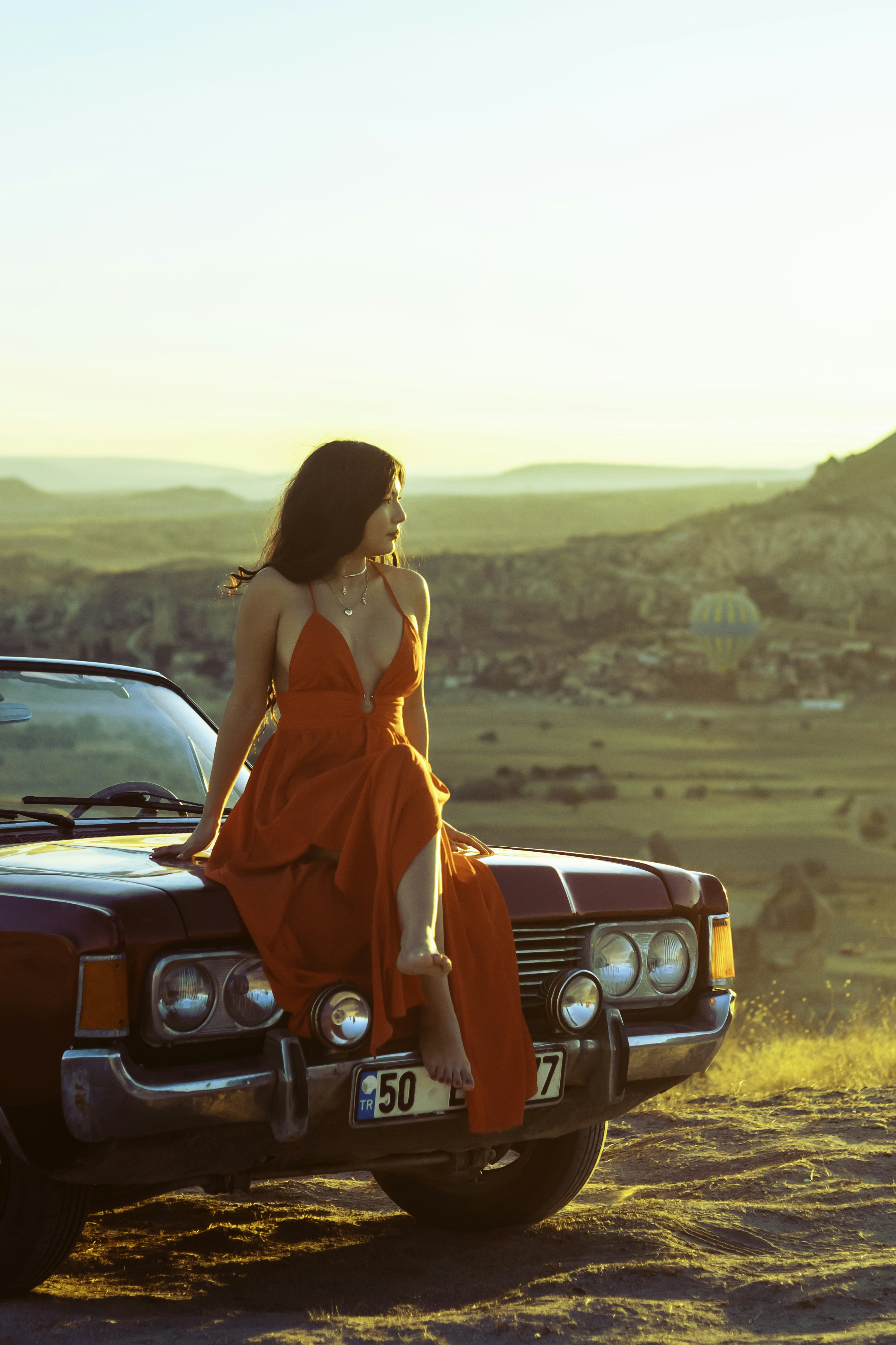 Red dress, dusty horizon: a woman perched on a vintage car at sunset