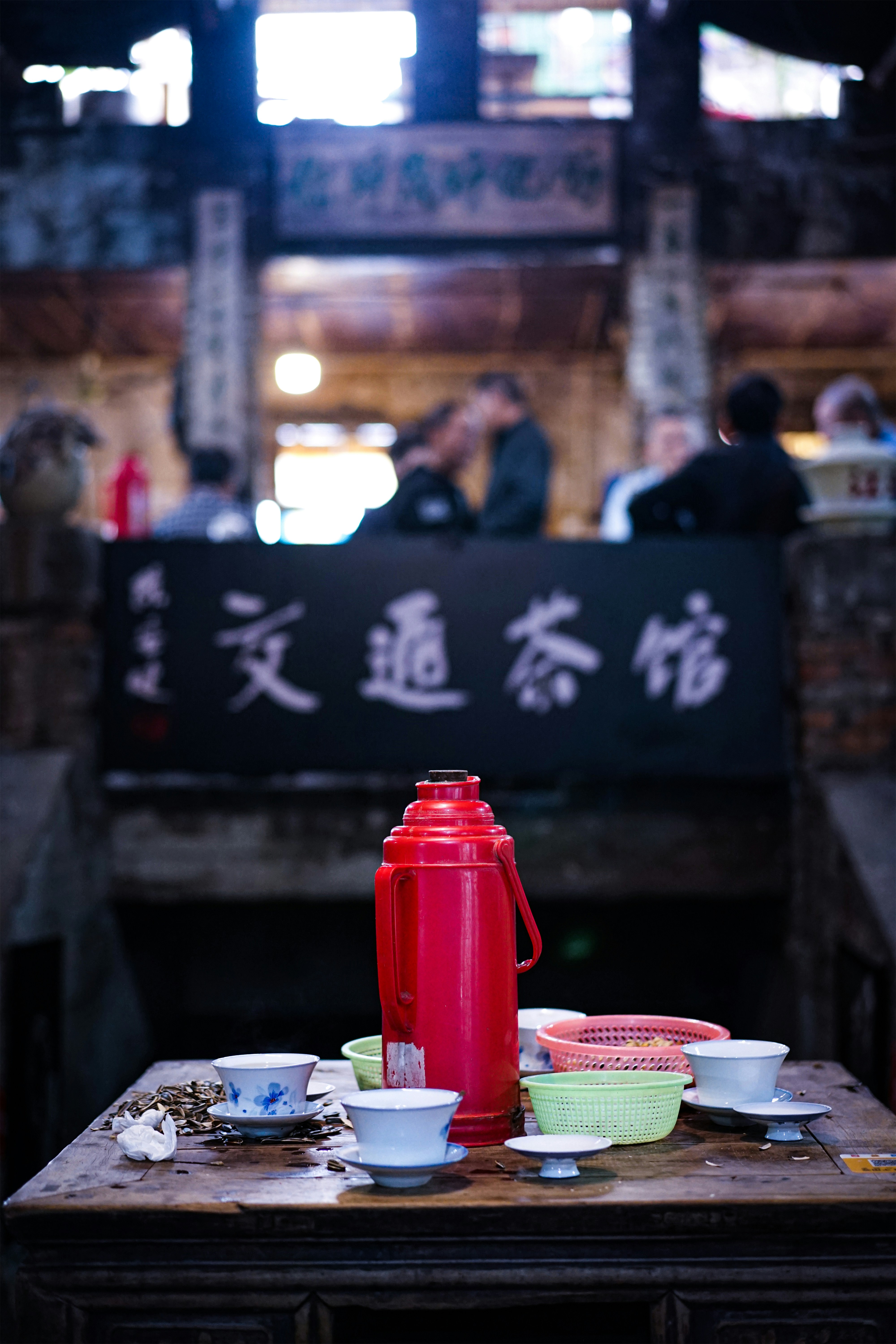 Vibrant red thermos and delicate tea cups arranged on a wooden table in a traditional tea house, with patrons engaged in conversation in the background.