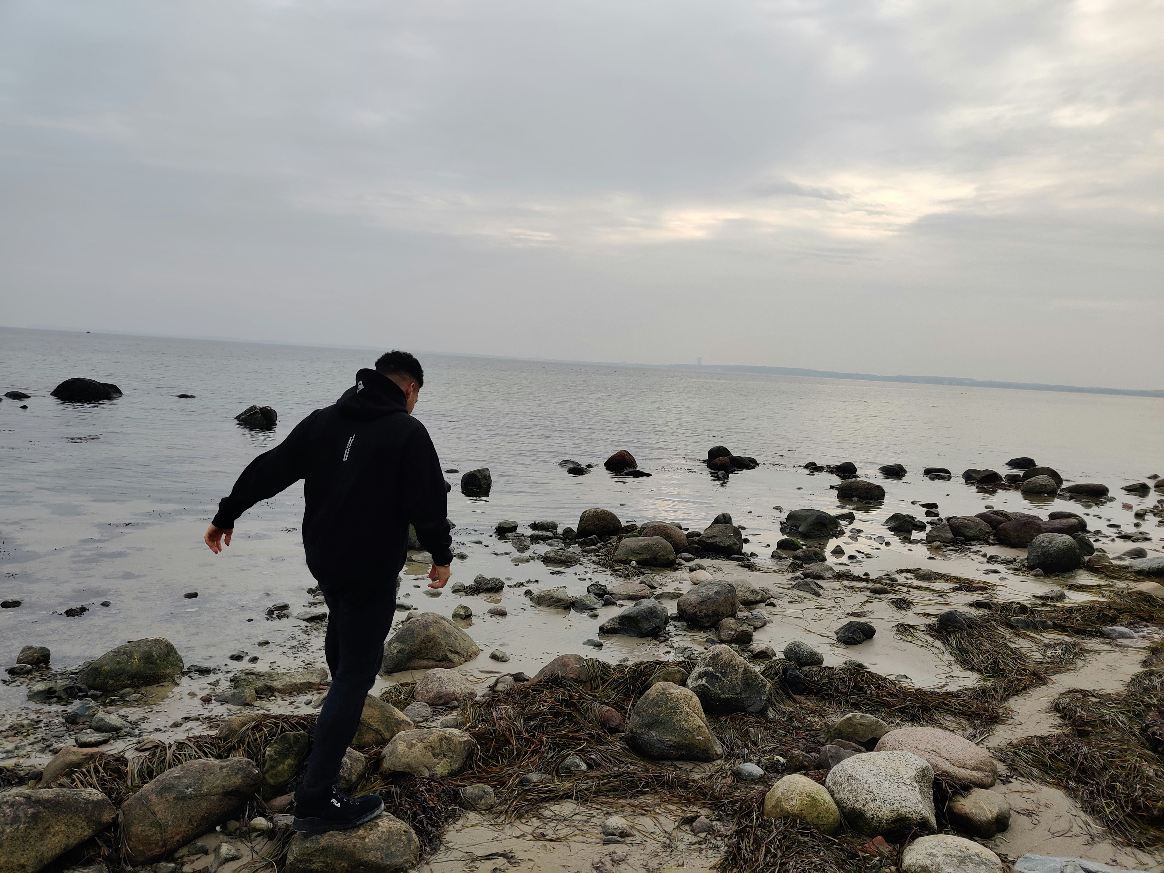A solitary figure in dark clothing walks along a rocky, tide-washed shoreline under a gray, overcast sky.