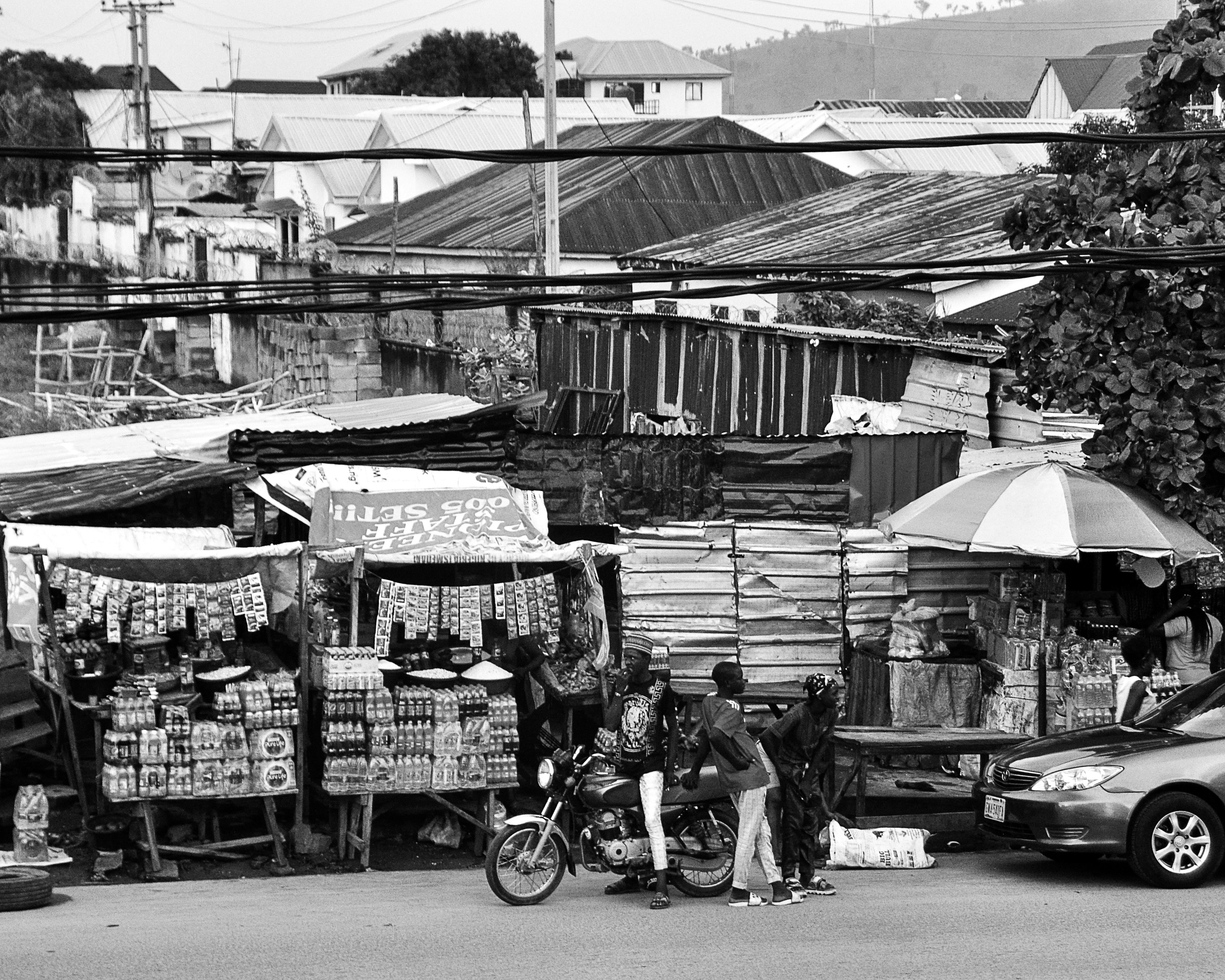 Busy street market scene with people gathered around a motorcycle amidst stalls and cars.