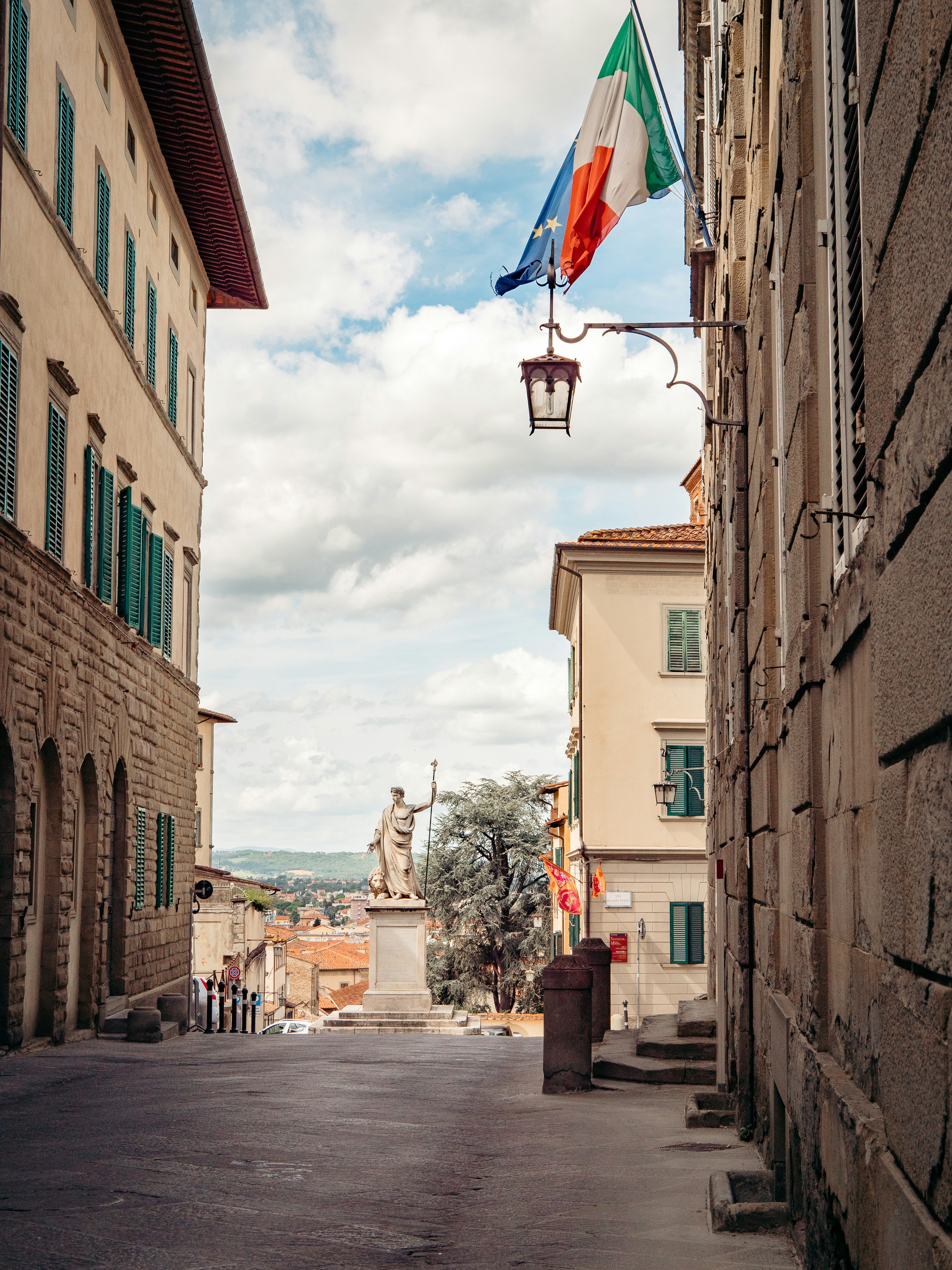 Flag over the quiet Tuscan street: a statue’s gaze down a sunlit avenue