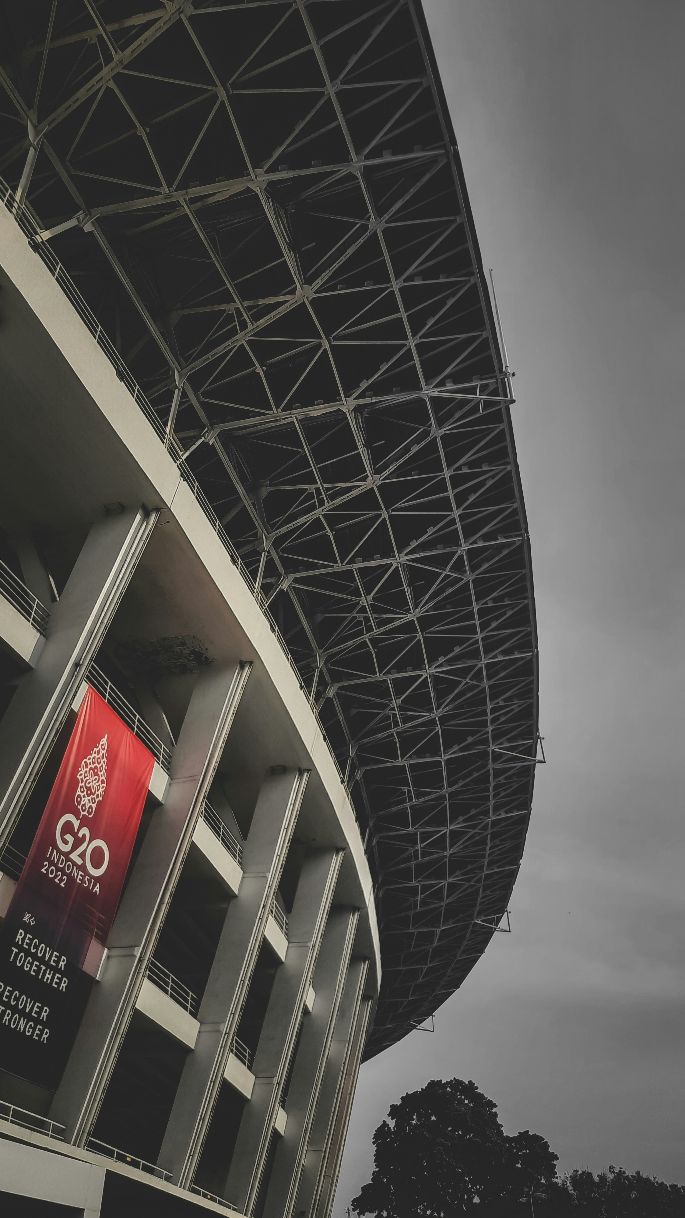 Architectural photograph of a sweeping stadium facade in grayscale, dominated by a lattice roof and a prominent vertical banner reading G20 Indonesia 2022. It emphasizes scale, rhythm, and industrial texture against a cloudy sky.