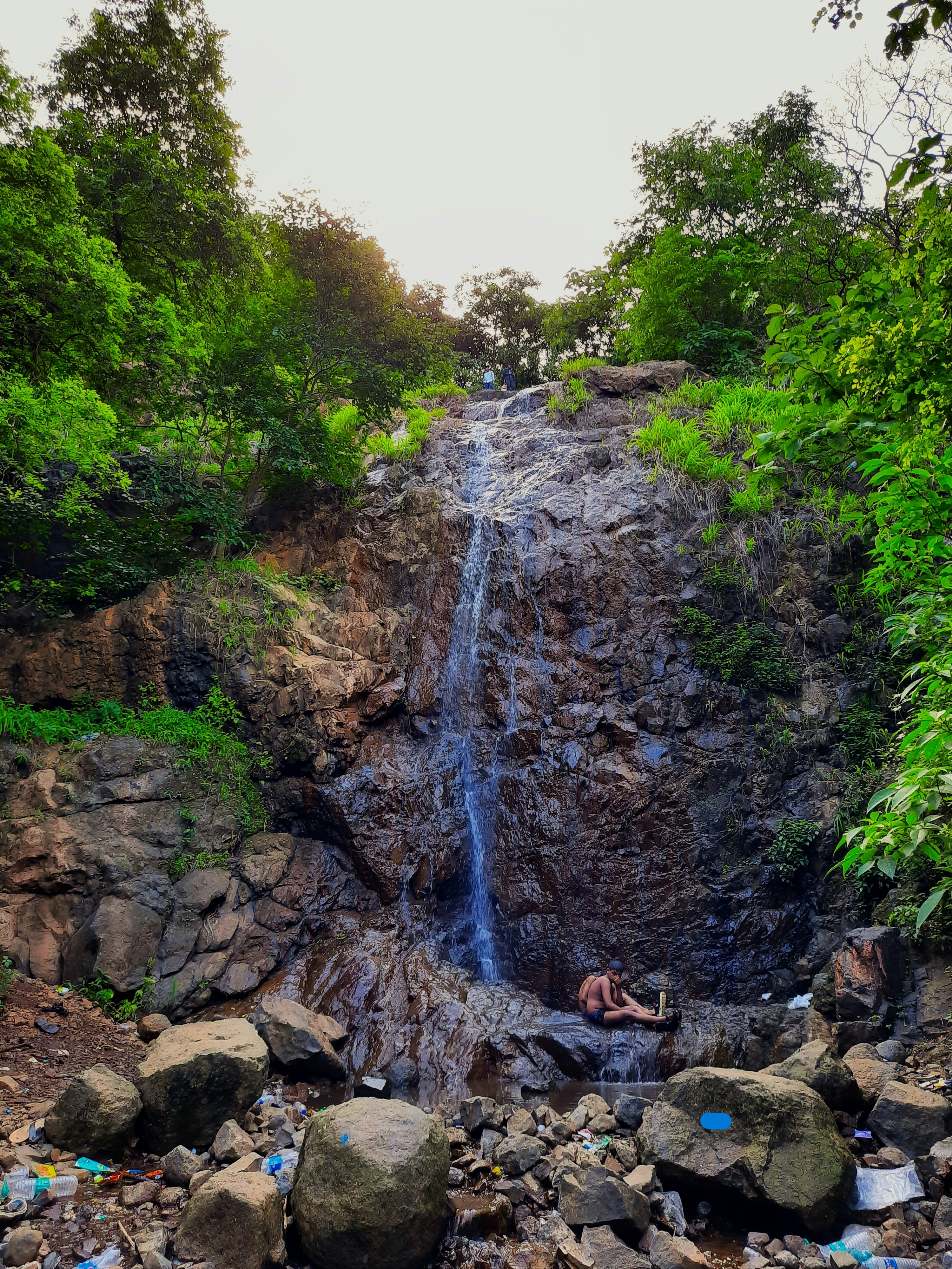 Photo of a small waterfall cascading down a rocky cliff, surrounded by lush trees; a person sits on rocks at the base.