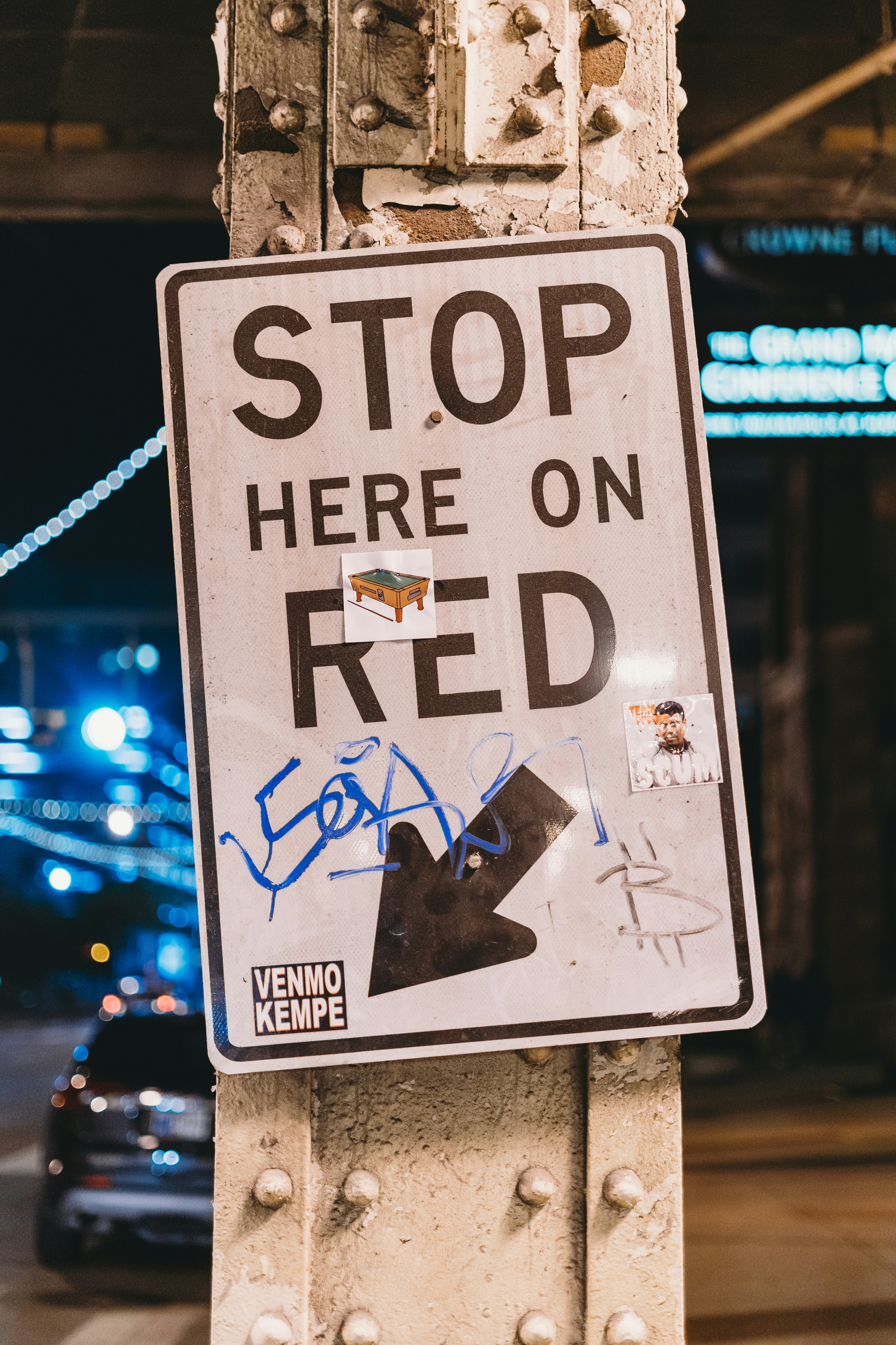 A weathered traffic sign with graffiti and stickers, set against a blurred urban backdrop at night.