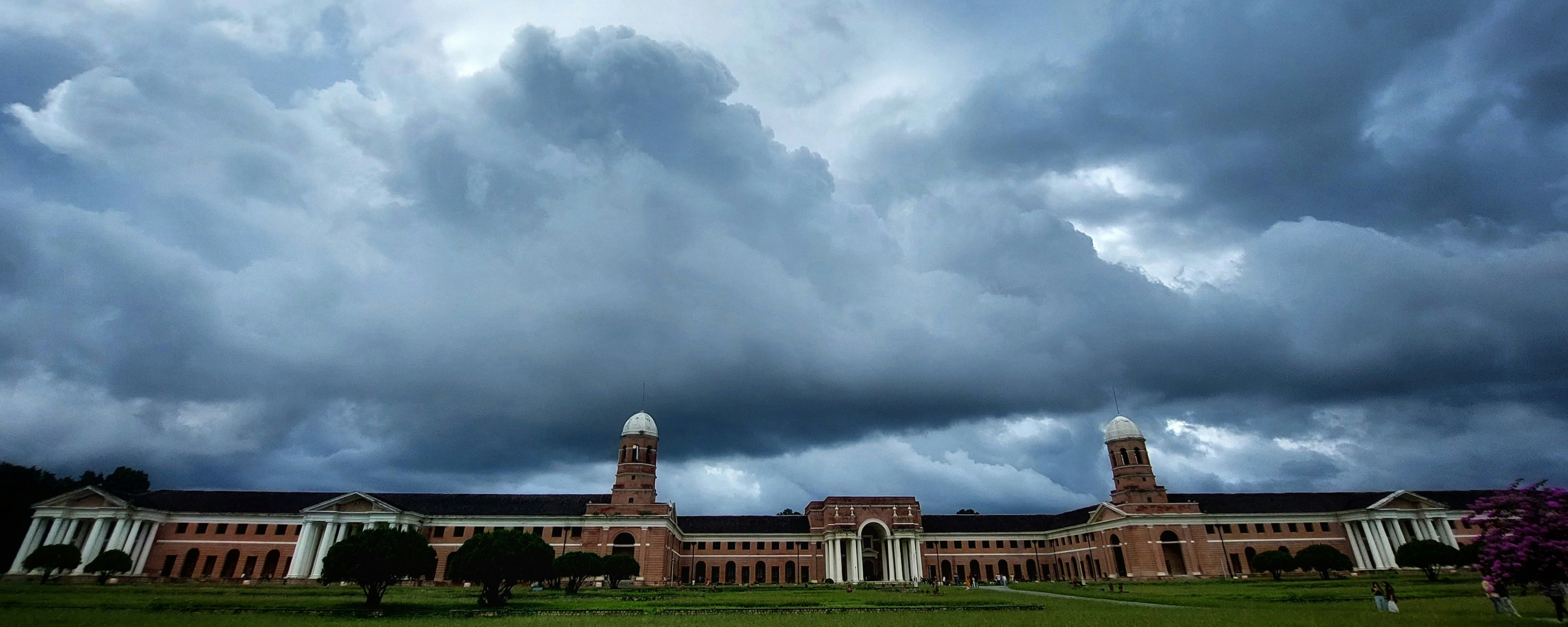 Dramatic storm clouds loom over a grand, historic brick building with towers and arches.