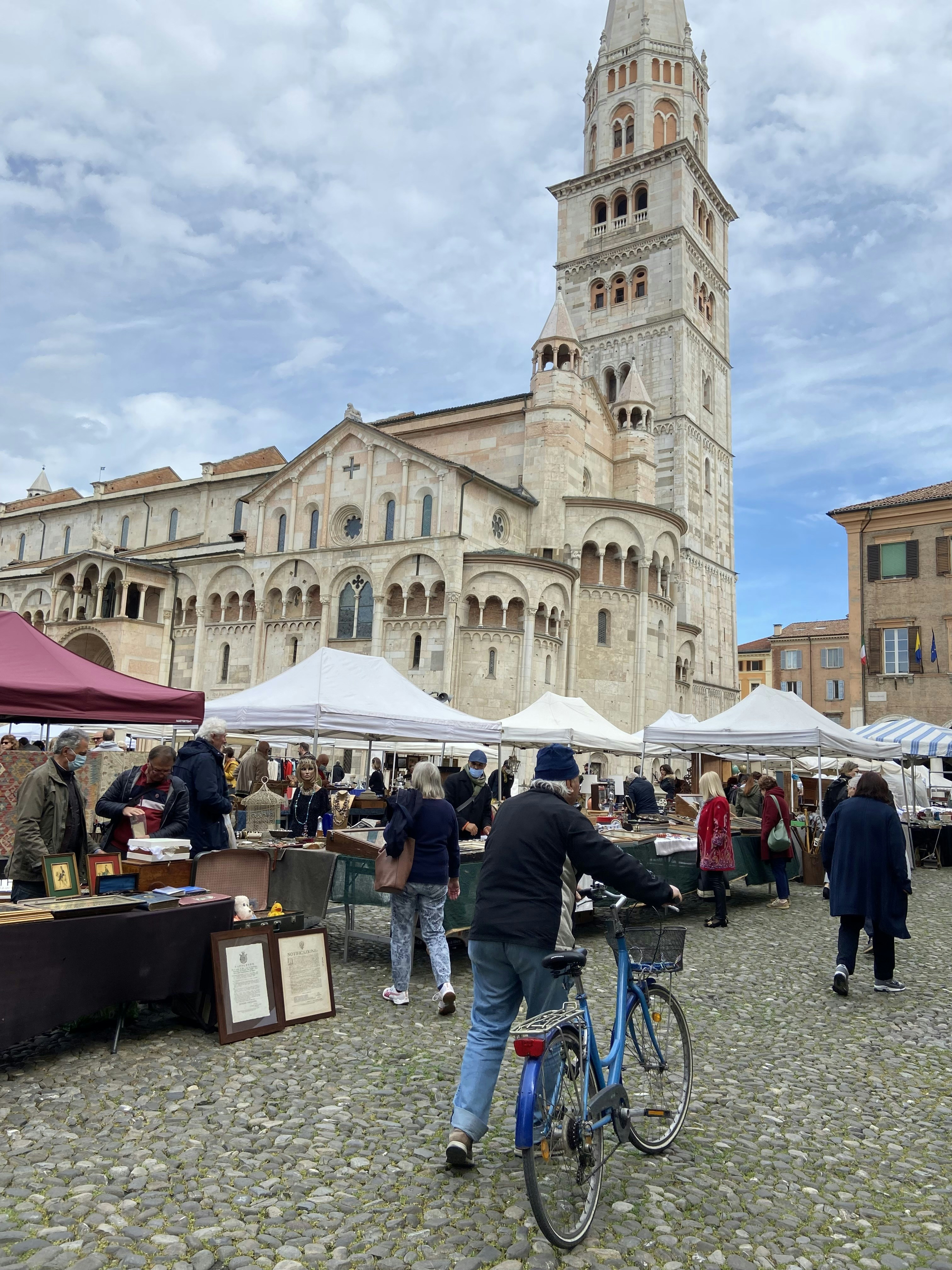 Bustling open-air market with vendors and visitors, set against a backdrop of an ancient church and bell tower.