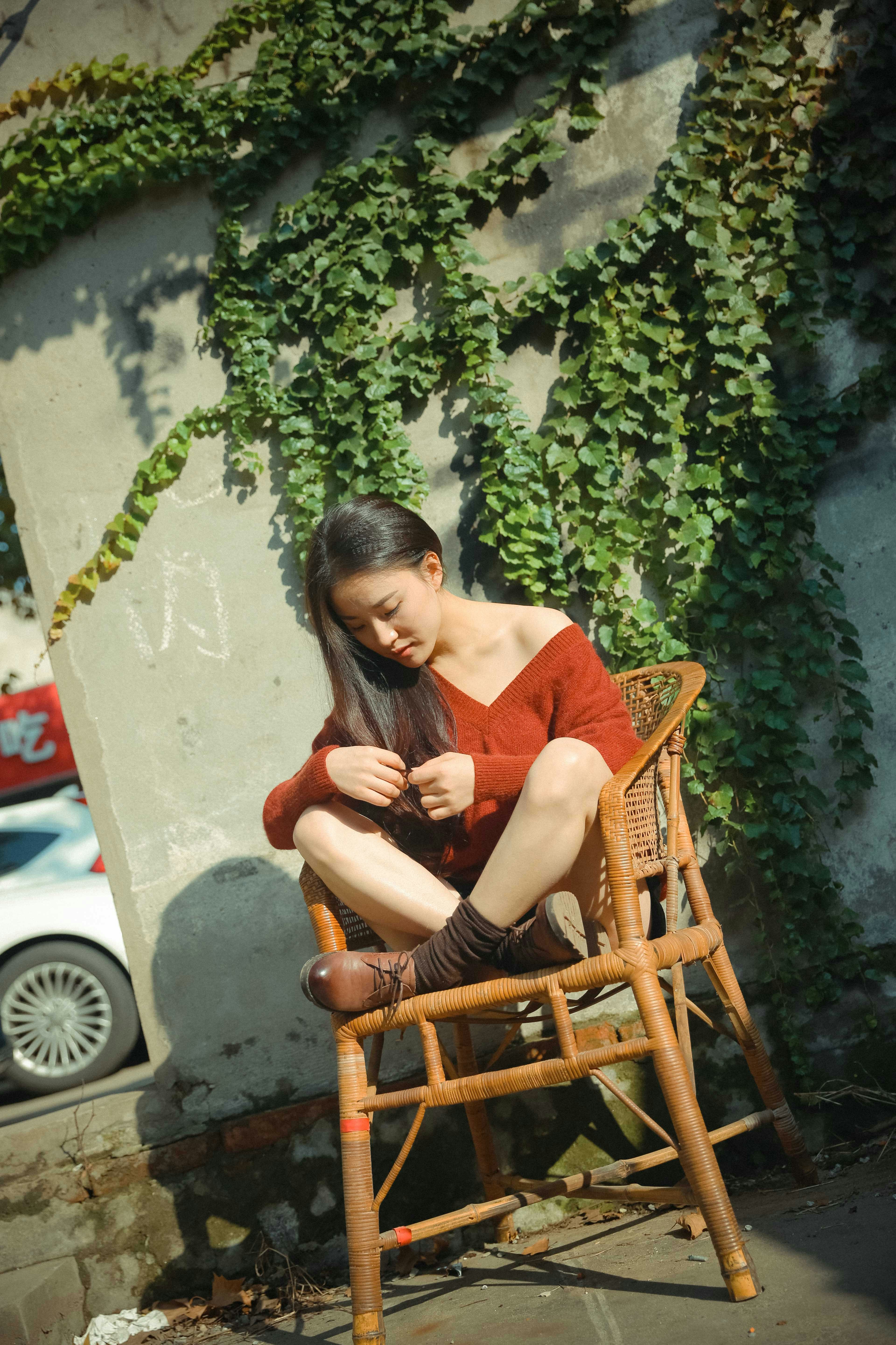A young woman in a cozy red sweater sits thoughtfully in a wicker chair surrounded by lush green ivy. The scene captures a serene moment in a natural setting.