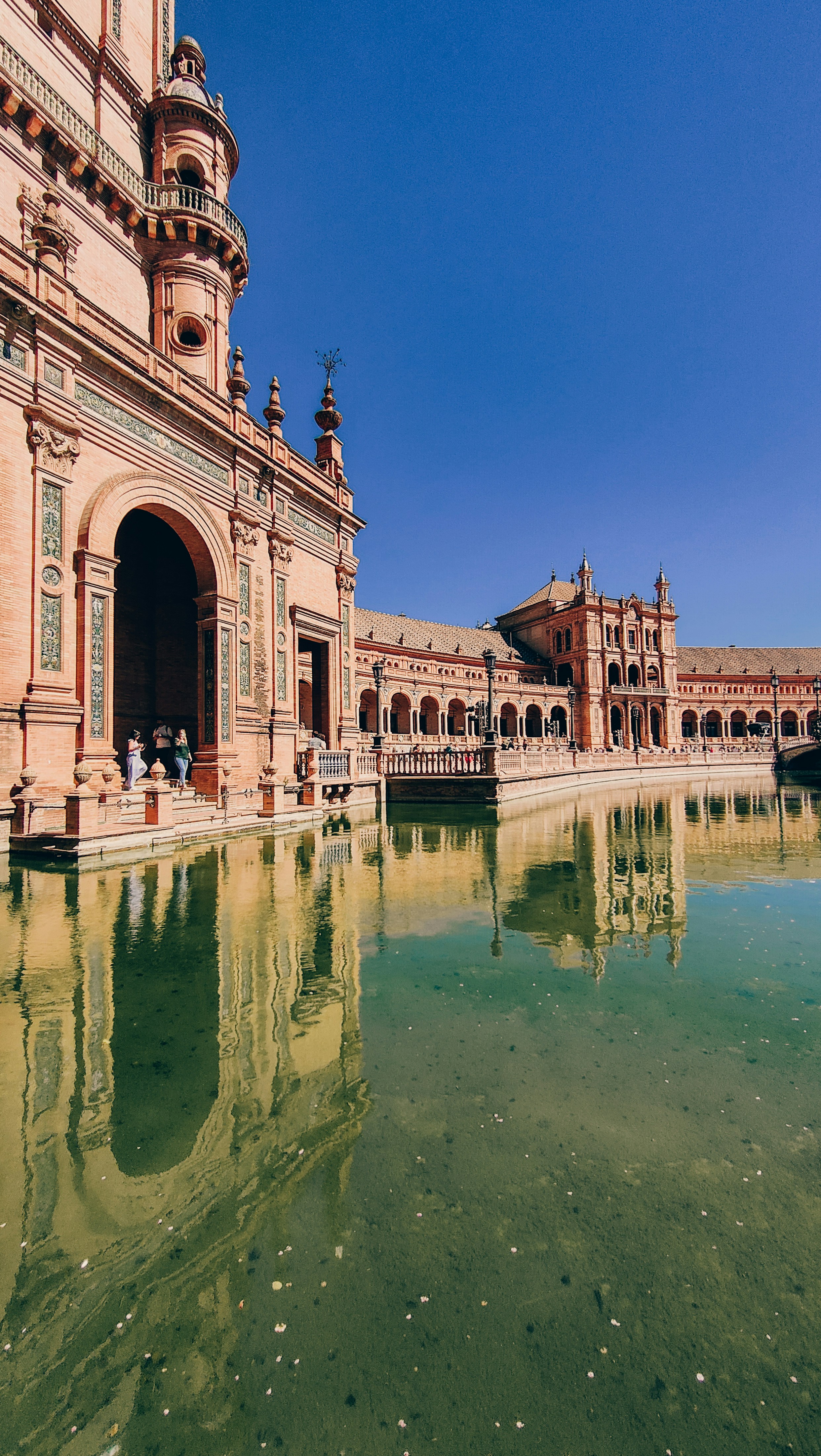Sunlit brick arches frame a calm canal at Plaza de España, Seville, with the ornate façade mirrored in the water.