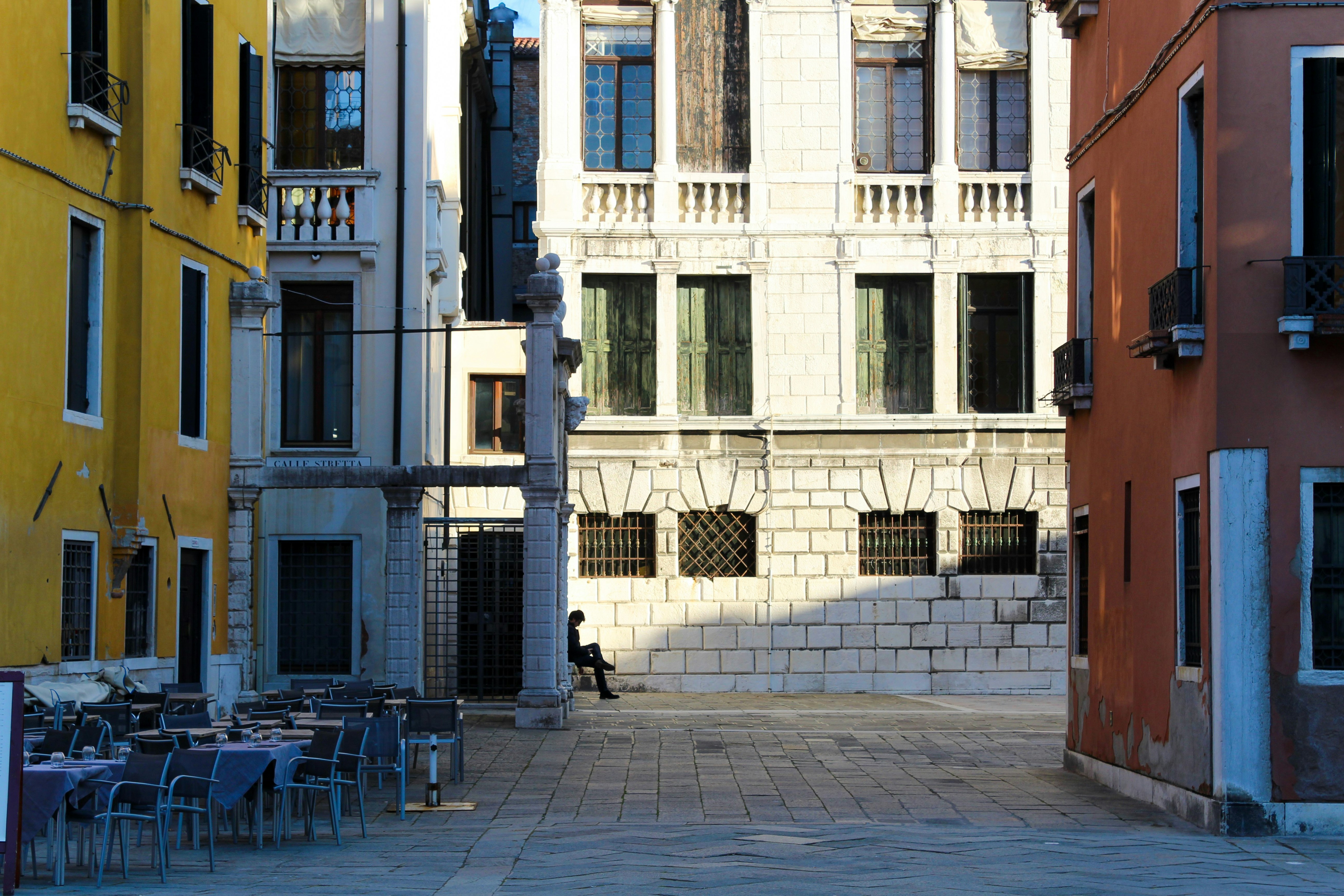 Solitary figure reading in a sunlit Venetian square, surrounded by colorful buildings and empty café tables.