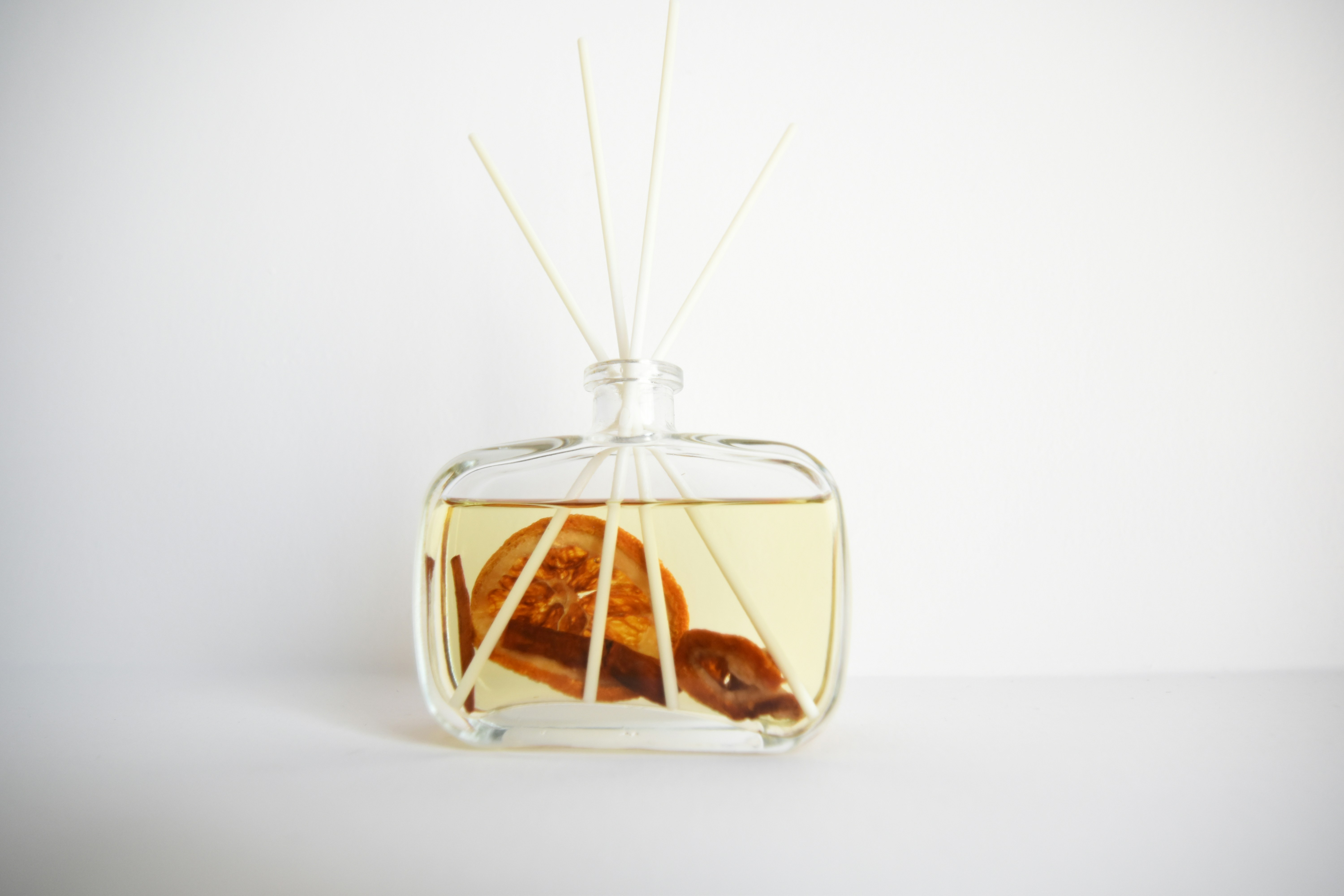 Close-up of a glass diffuser bottle with white reeds and pale liquid. A dried citrus slice is visible inside, set against a clean white background.