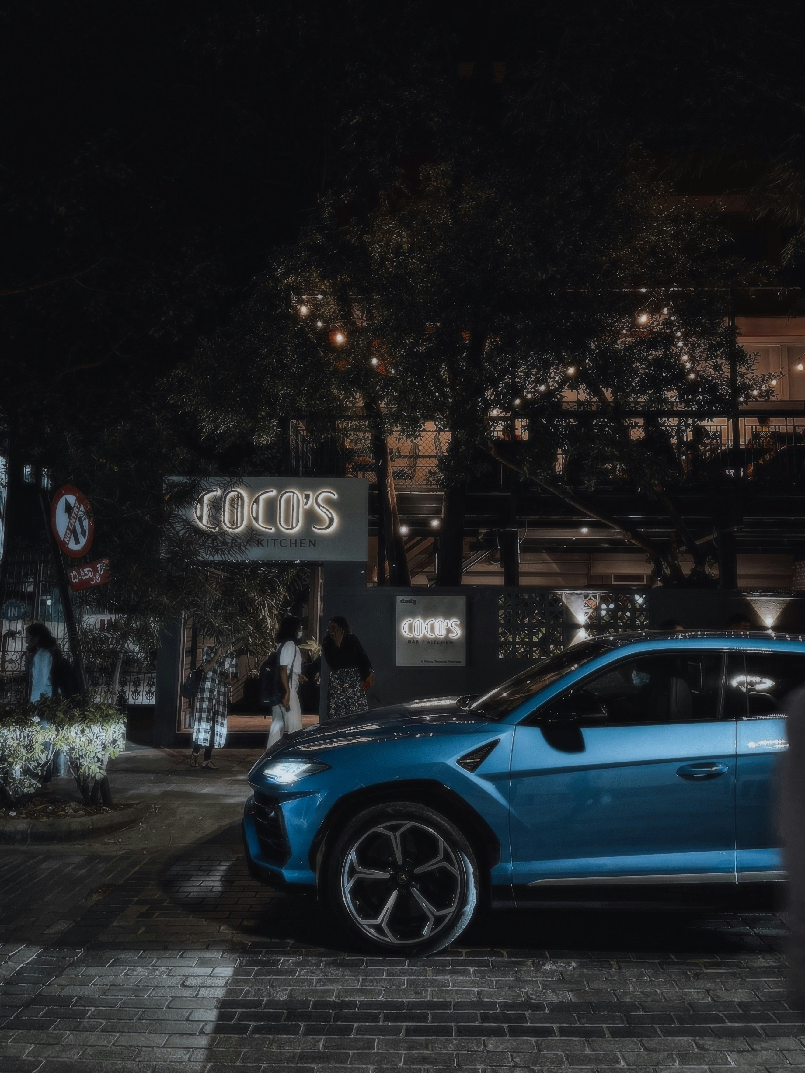 A sleek blue SUV parked outside Coco's Kitchen, illuminated by ambient lights. The bustling atmosphere is highlighted by people mingling near the entrance.