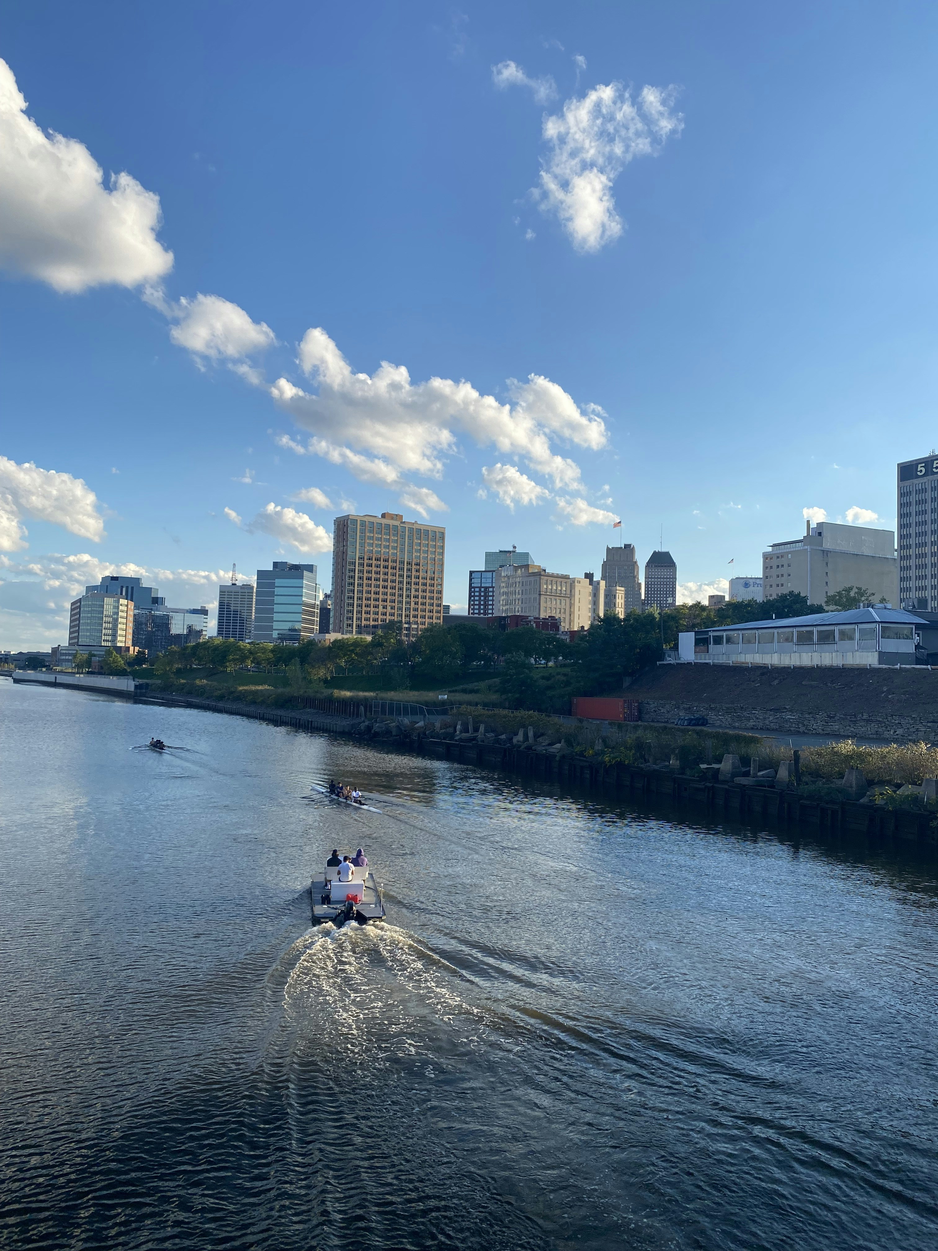 Boats glide along a tranquil river reflecting the skyline of a bustling city under a bright blue sky with scattered clouds.