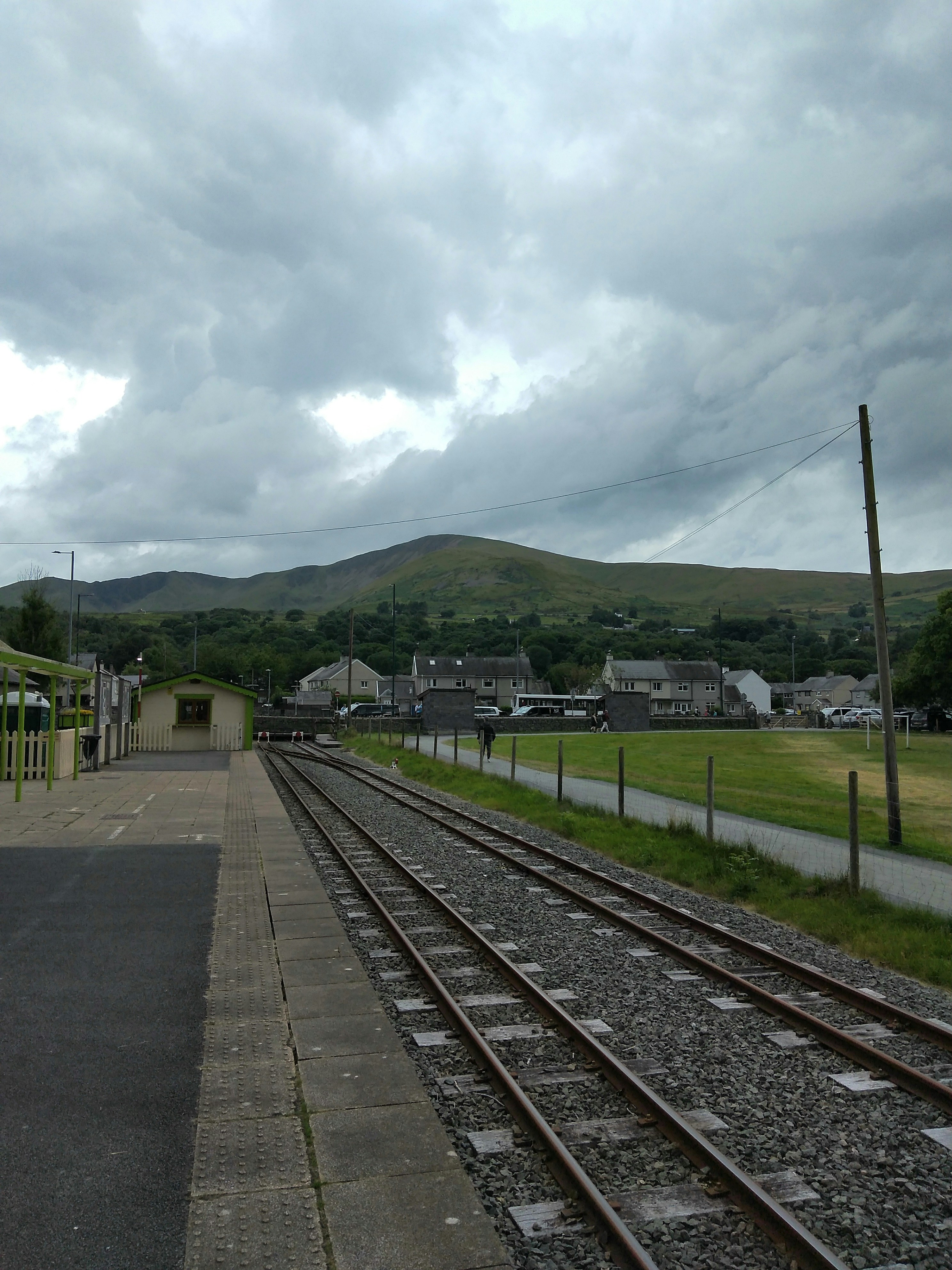A tranquil railway station scene nestled between rolling hills and quaint village houses under a moody sky.