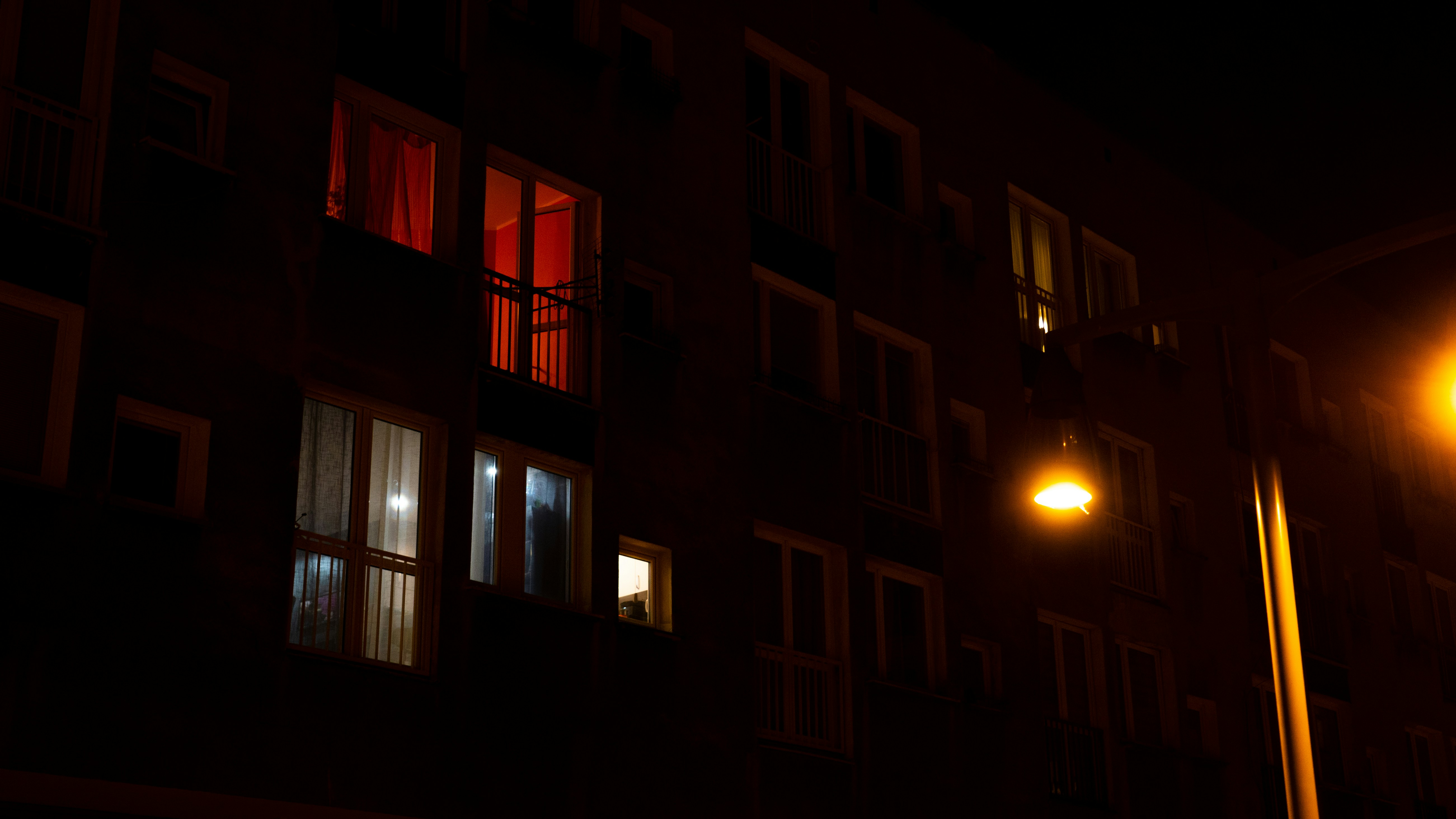 Illuminated apartment windows reveal glimpses of life inside a darkened building at night, contrasting warm light against the cool shadows outside.