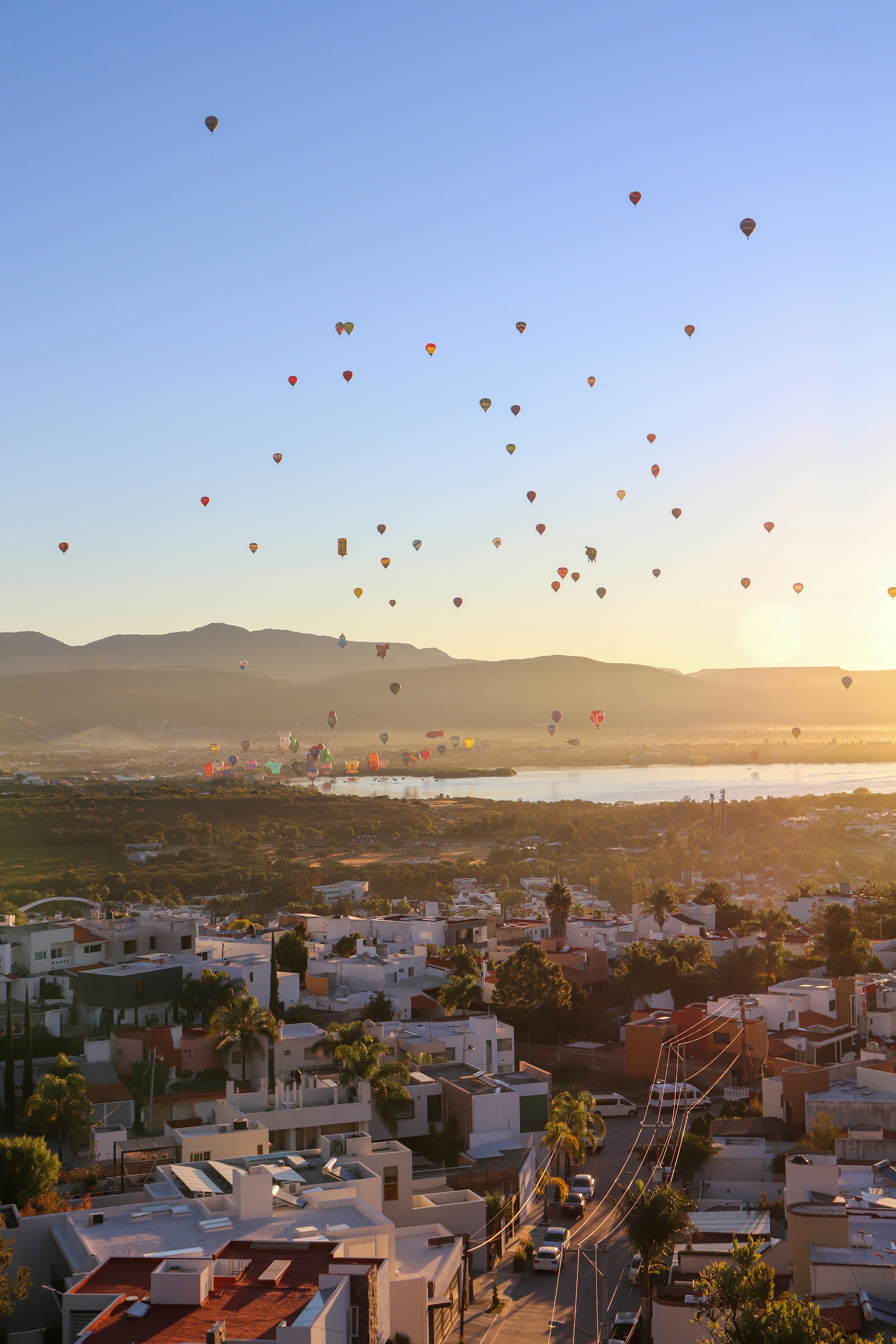 Hot air balloons rising above a cityscape at sunrise with distant mountains and a serene river.