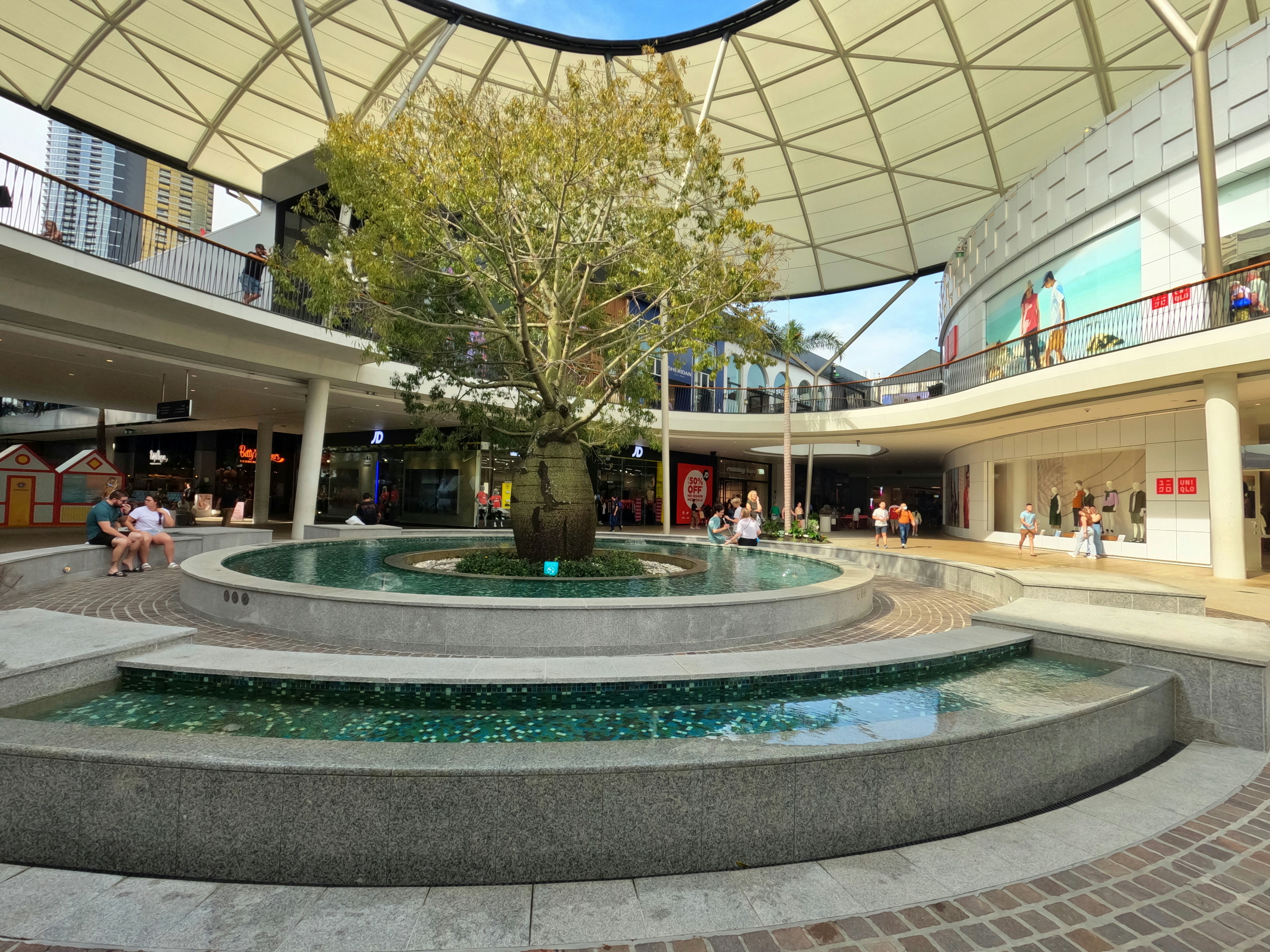 A central water feature with a large tree surrounded by tiered stone platforms in a modern shopping complex. Visitors relax nearby, enjoying the tranquil atmosphere.