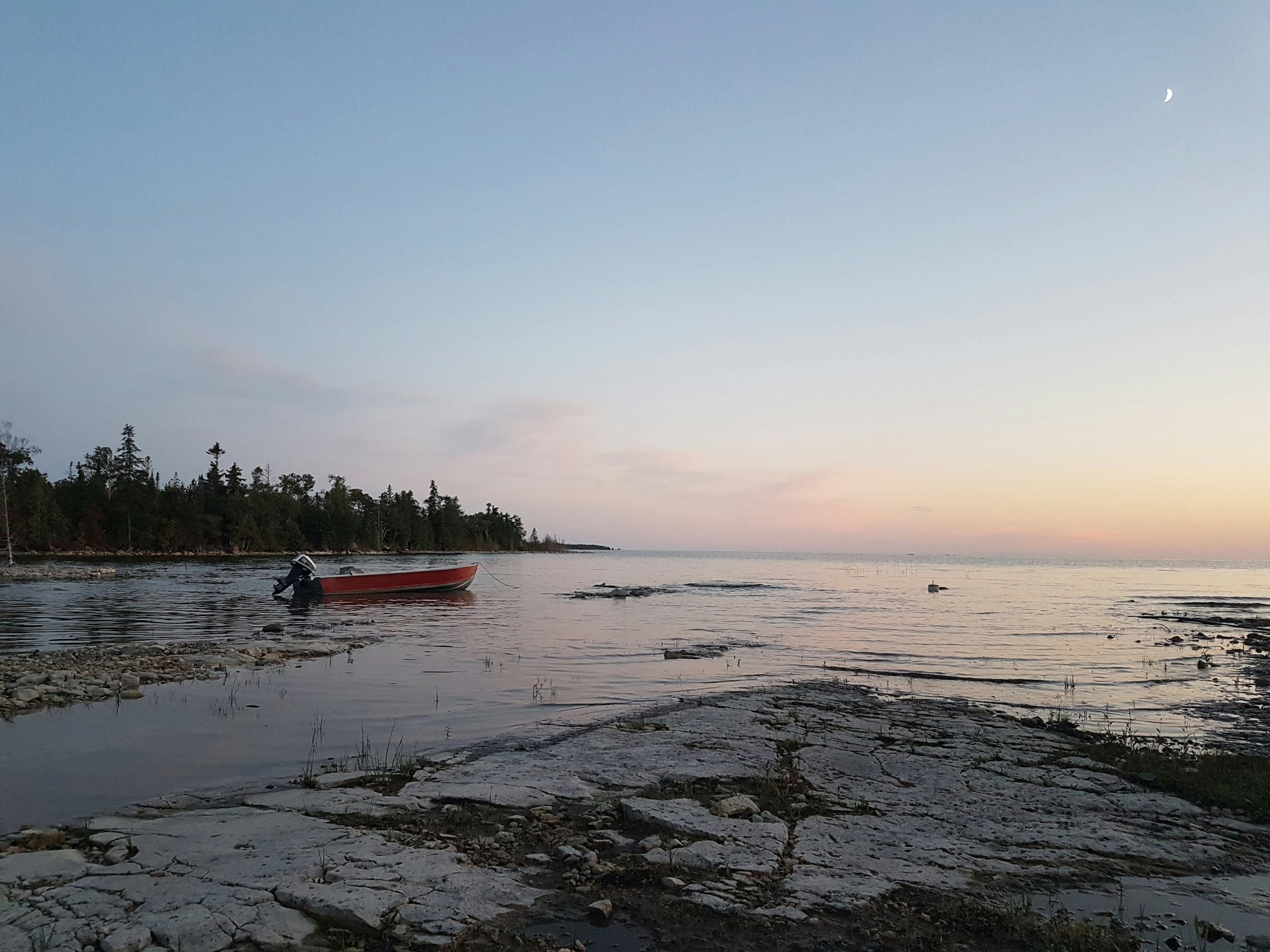 A red boat anchored near rocky shores under a pastel sky at dusk. The calm water reflects the fading light of day.