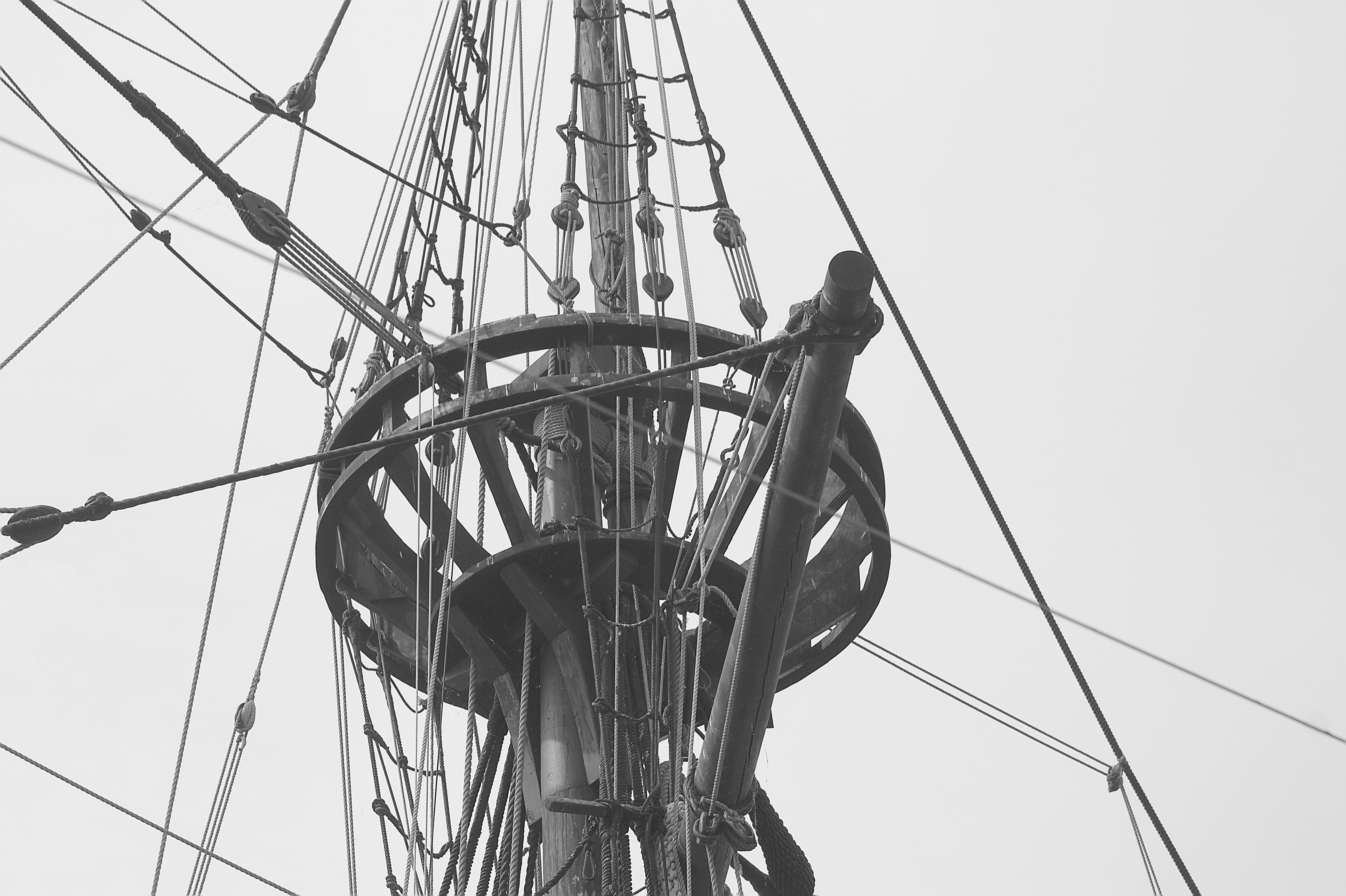 Black and white photograph of a ship's mast with intricate rigging and crow's nest.
