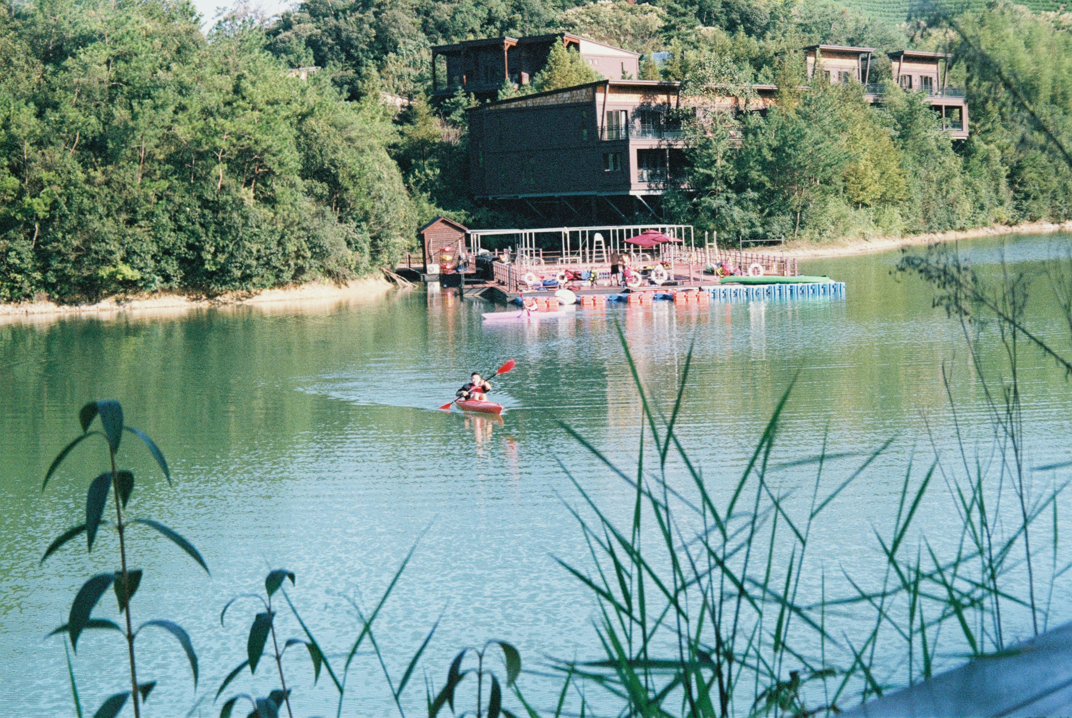 Kayaker glides across a calm lake surrounded by lush greenery and rustic buildings.