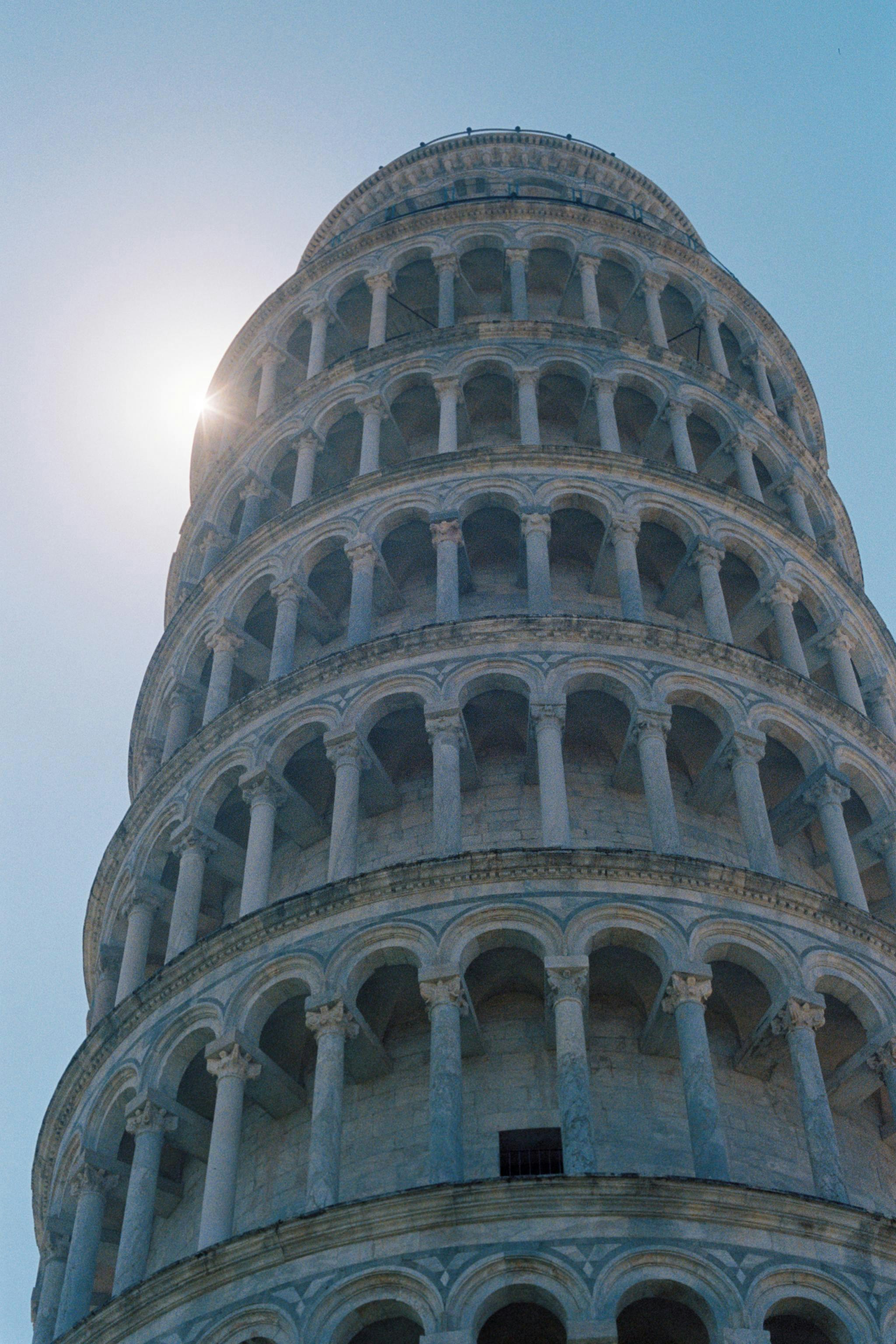 Sunlit arches encircle the Leaning Tower of Pisa as sunlight streams from the left, casting a subtle halo and lens flare over the stone facade.